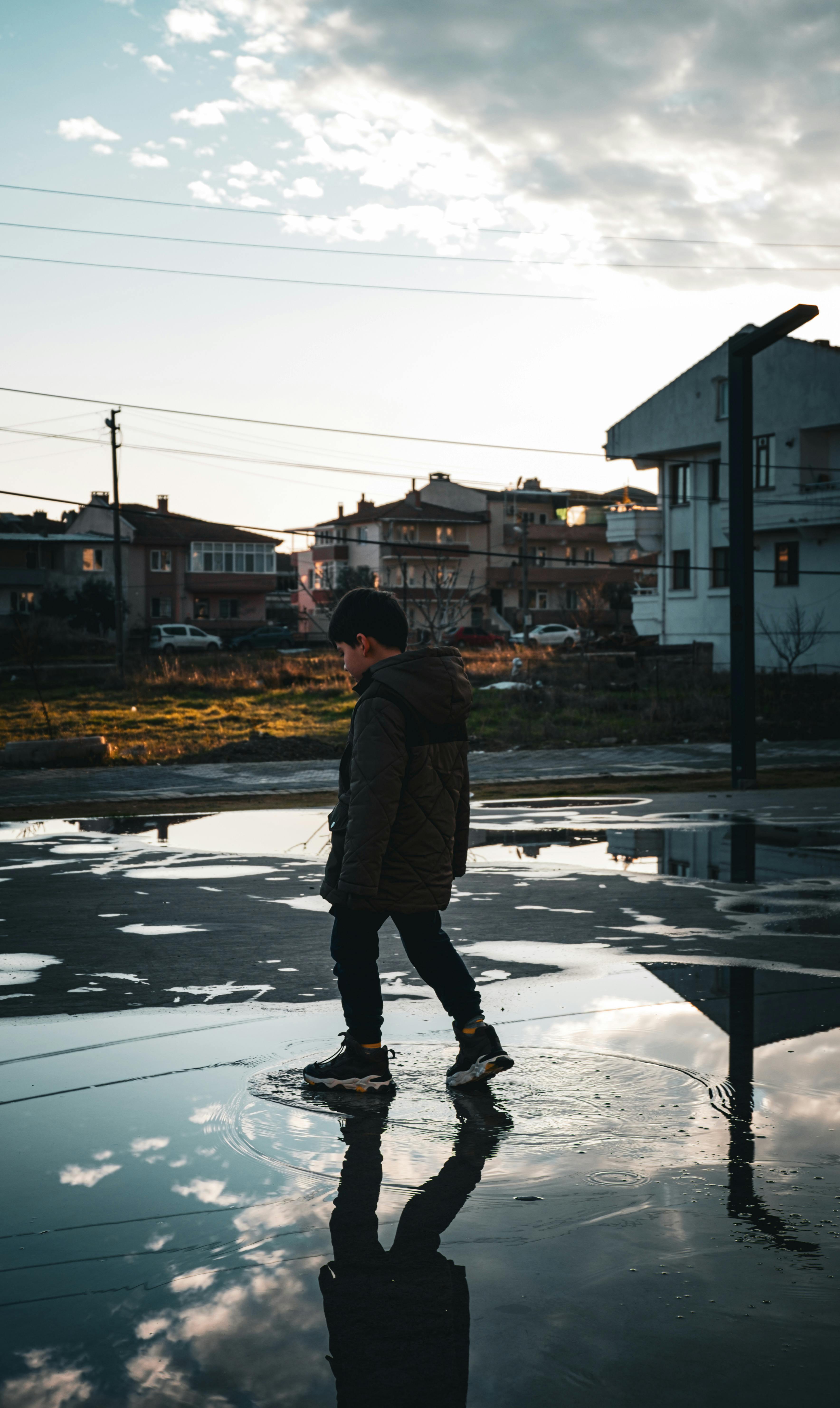 Young Boy Walking in Urban Puddle Scene at Dusk · Free Stock Photo