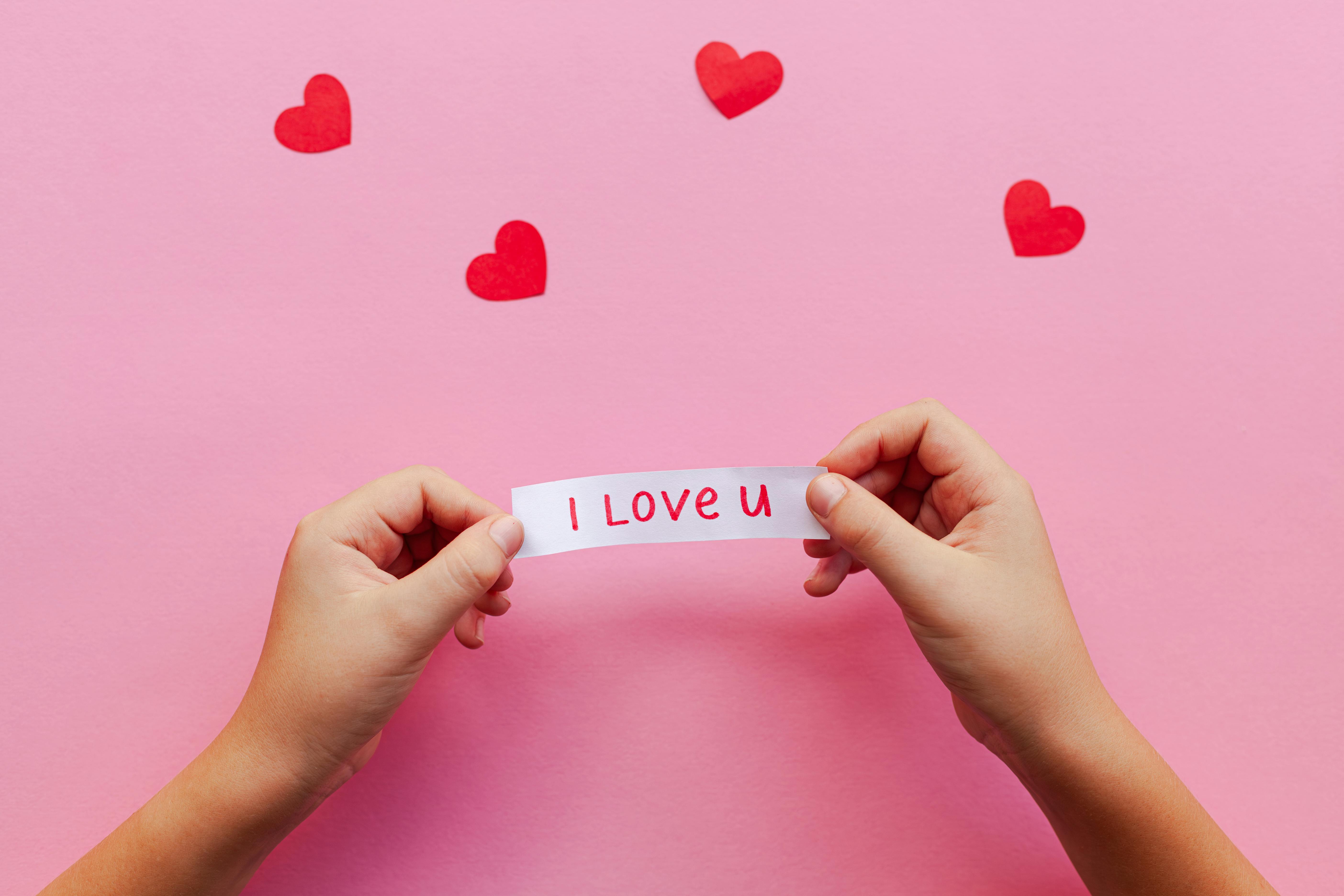 Hands holding 'I Love U' note on pink background · Free Stock Photo