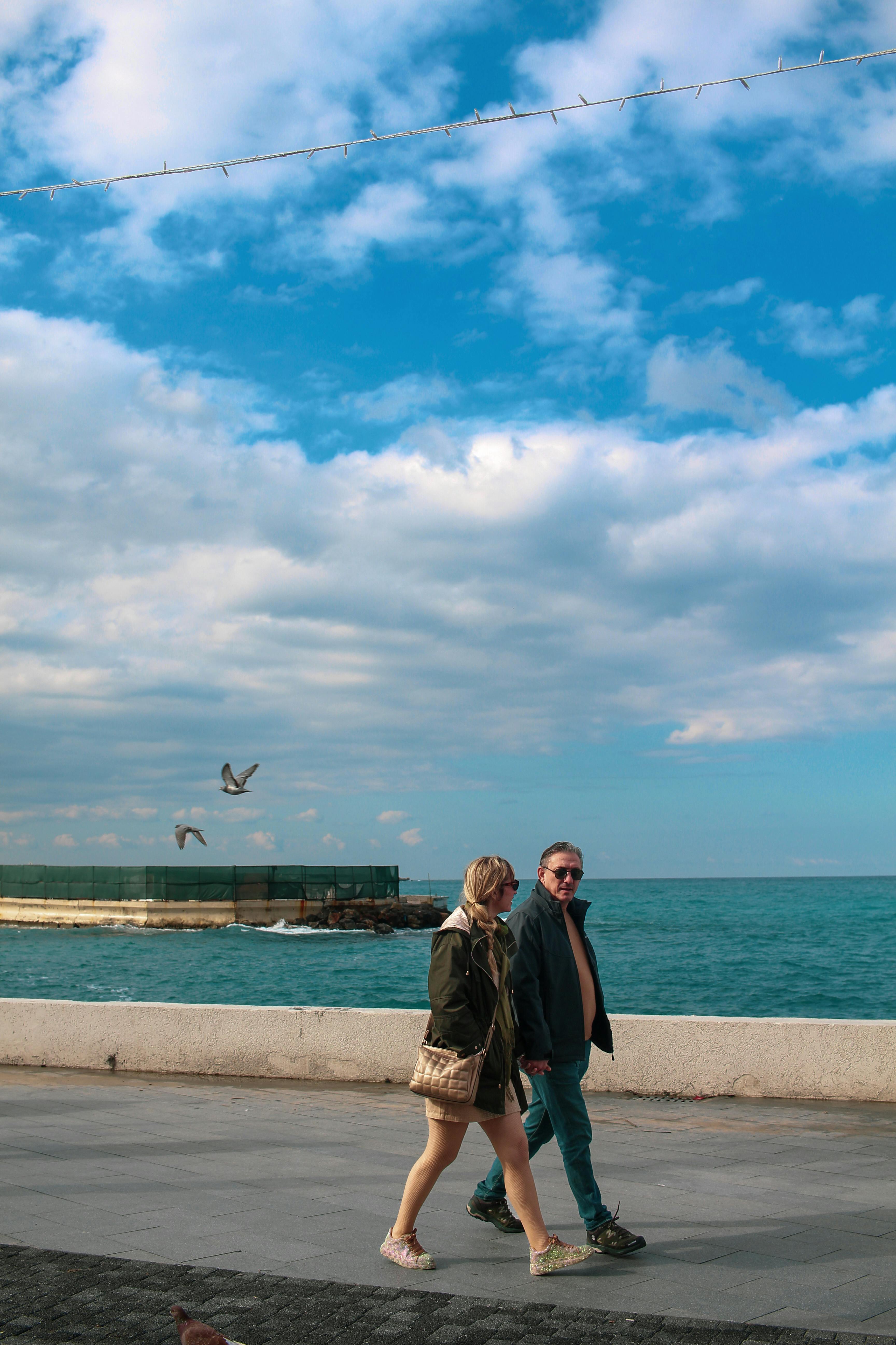 Couple Enjoying a Seaside Stroll Under Blue Skies · Free Stock Photo