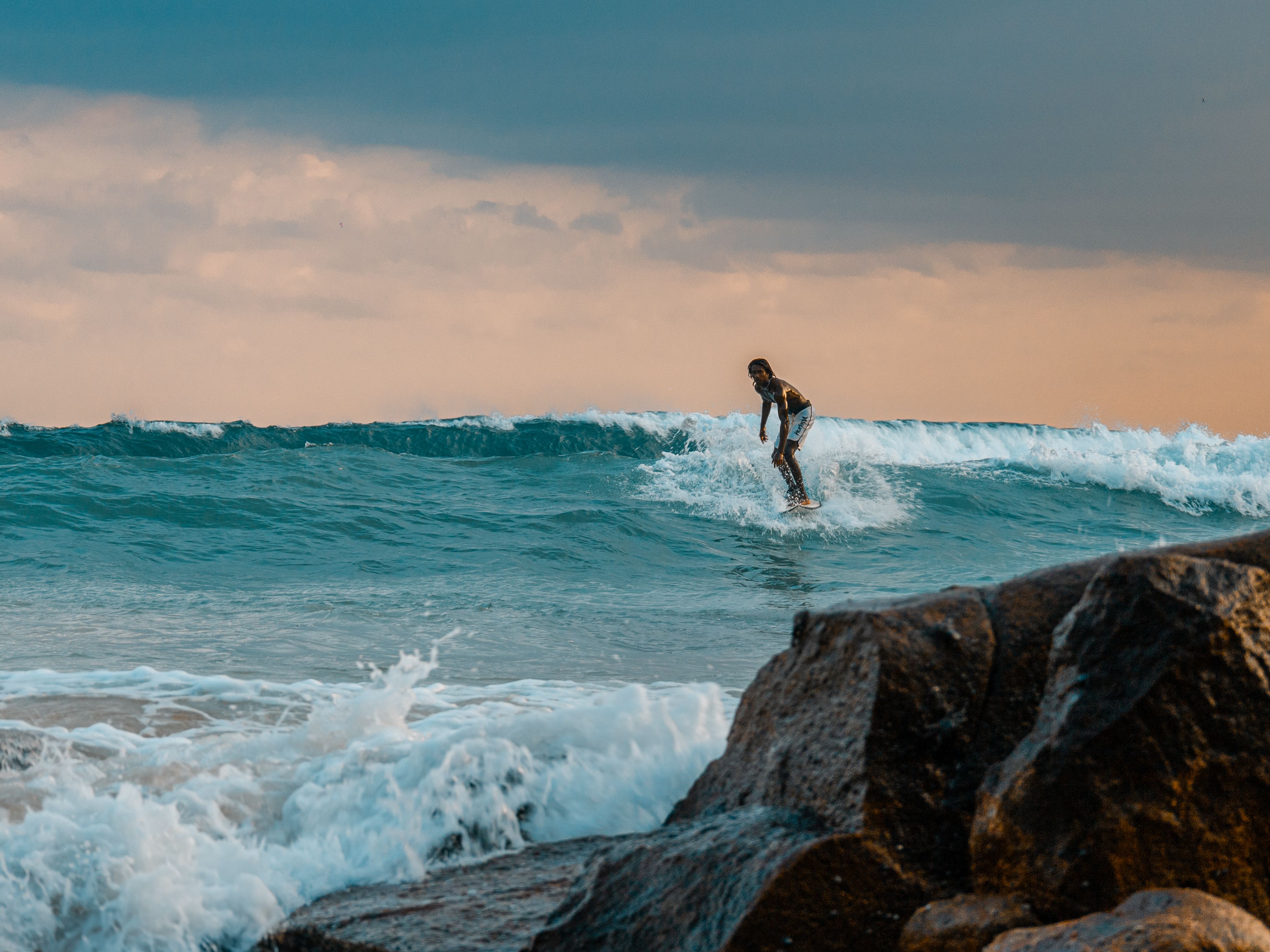 Surfer riding waves near rocky shore at sunset · Free Stock Photo