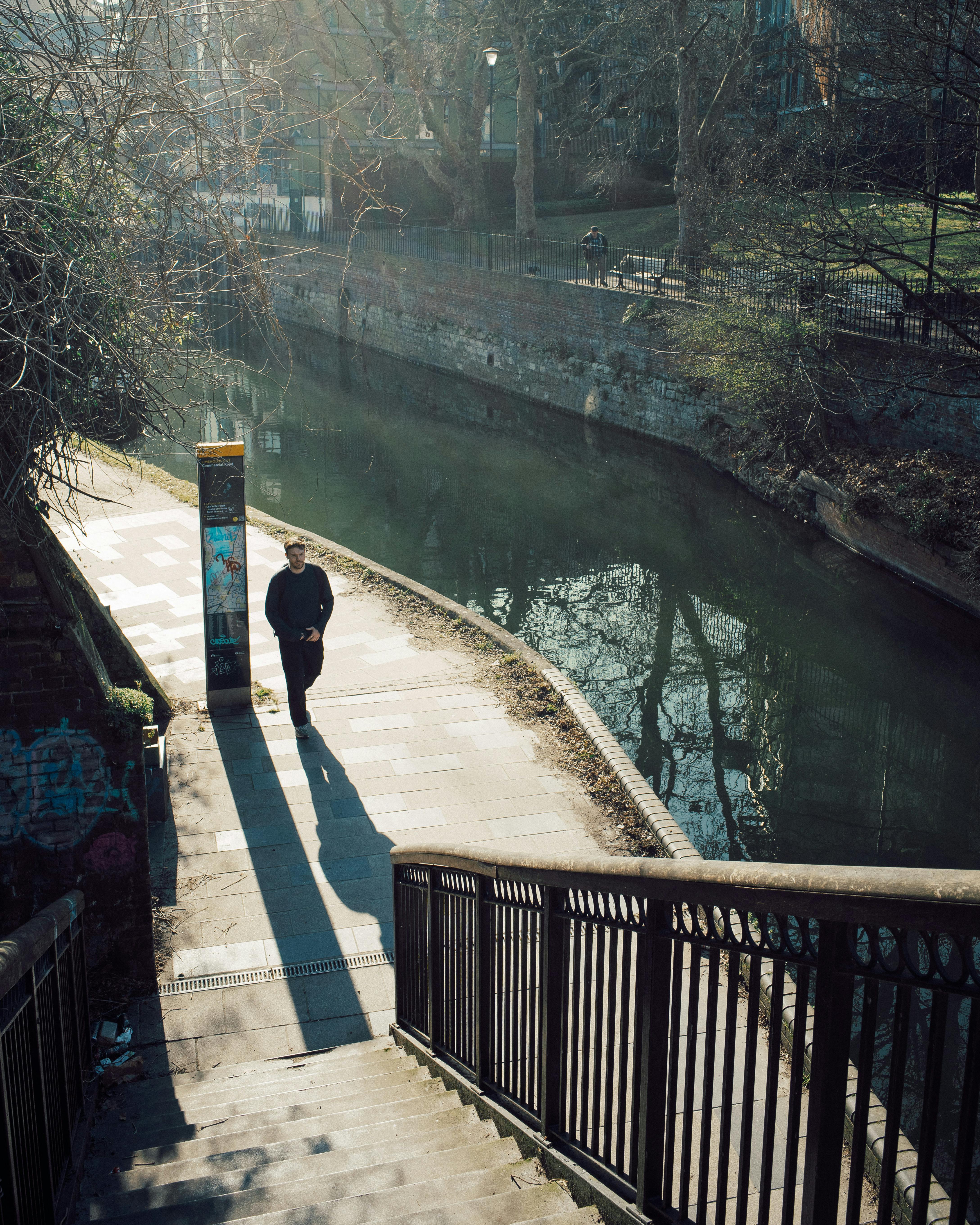 Man Walking Along Riverside Pathway in City Park · Free Stock Photo