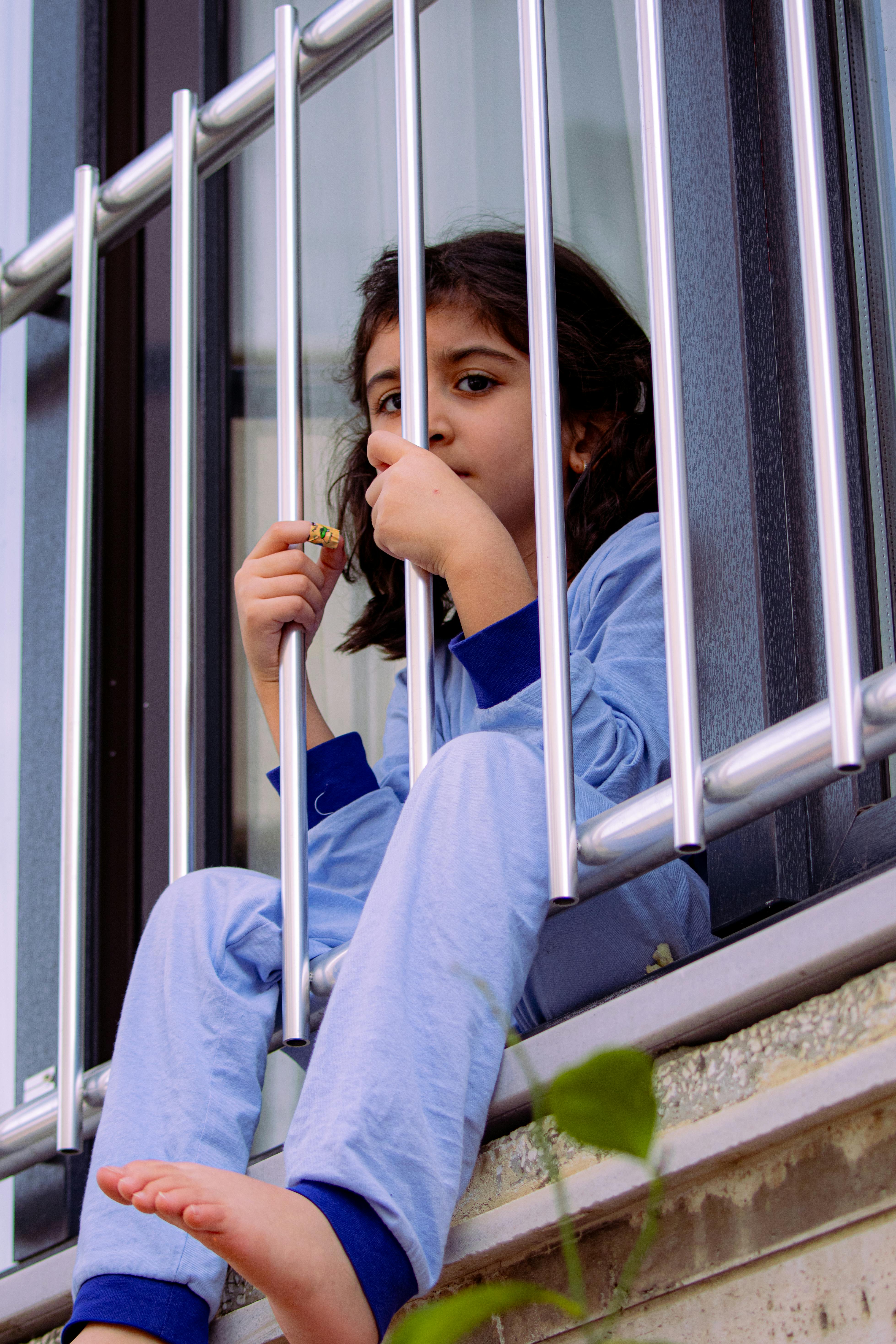 Child Sitting in Window Grating in Urban Setting · Free Stock Photo
