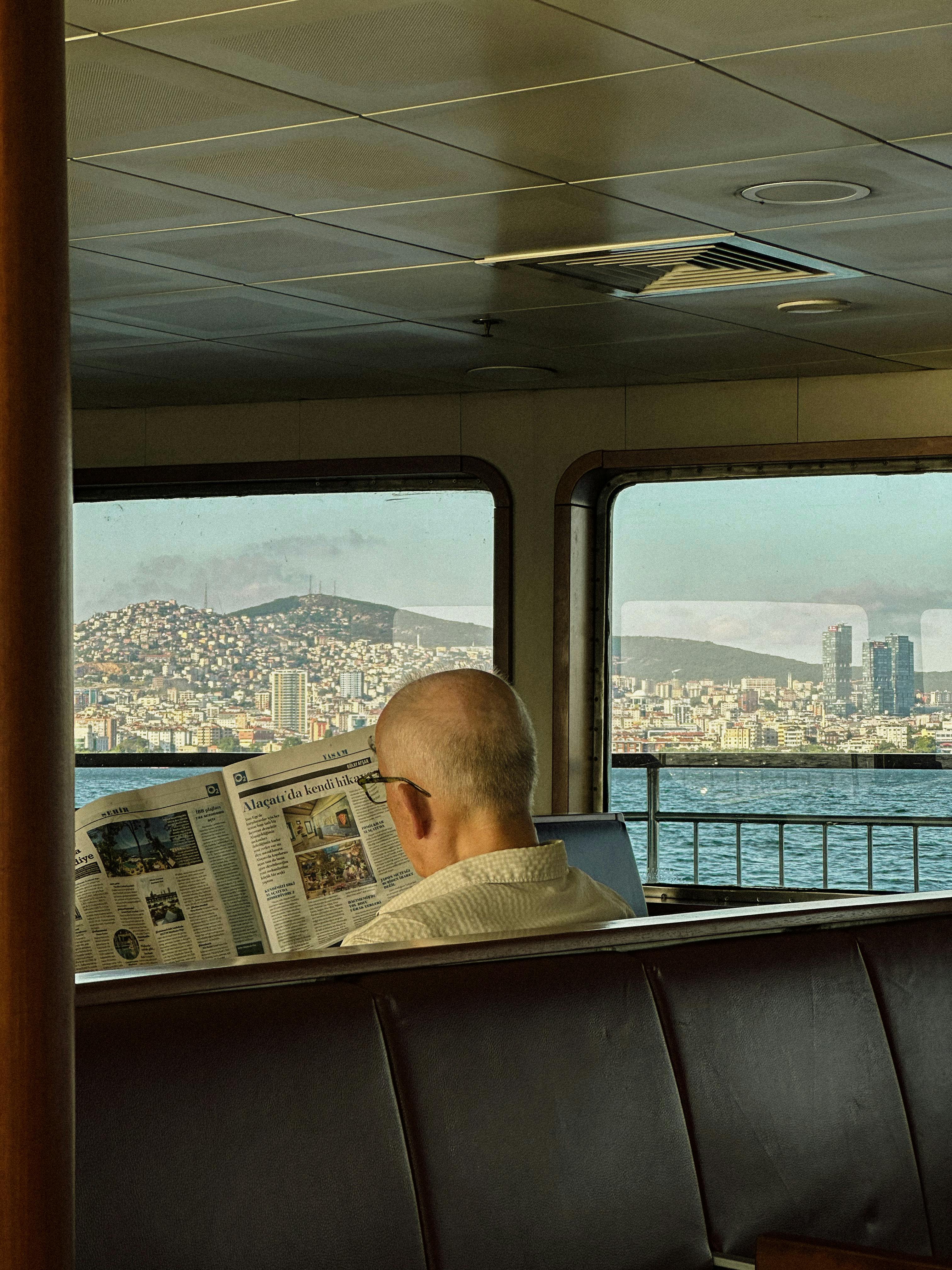 Man Reading Newspaper on Ferry Overlooking Cityscape · Free Stock Photo
