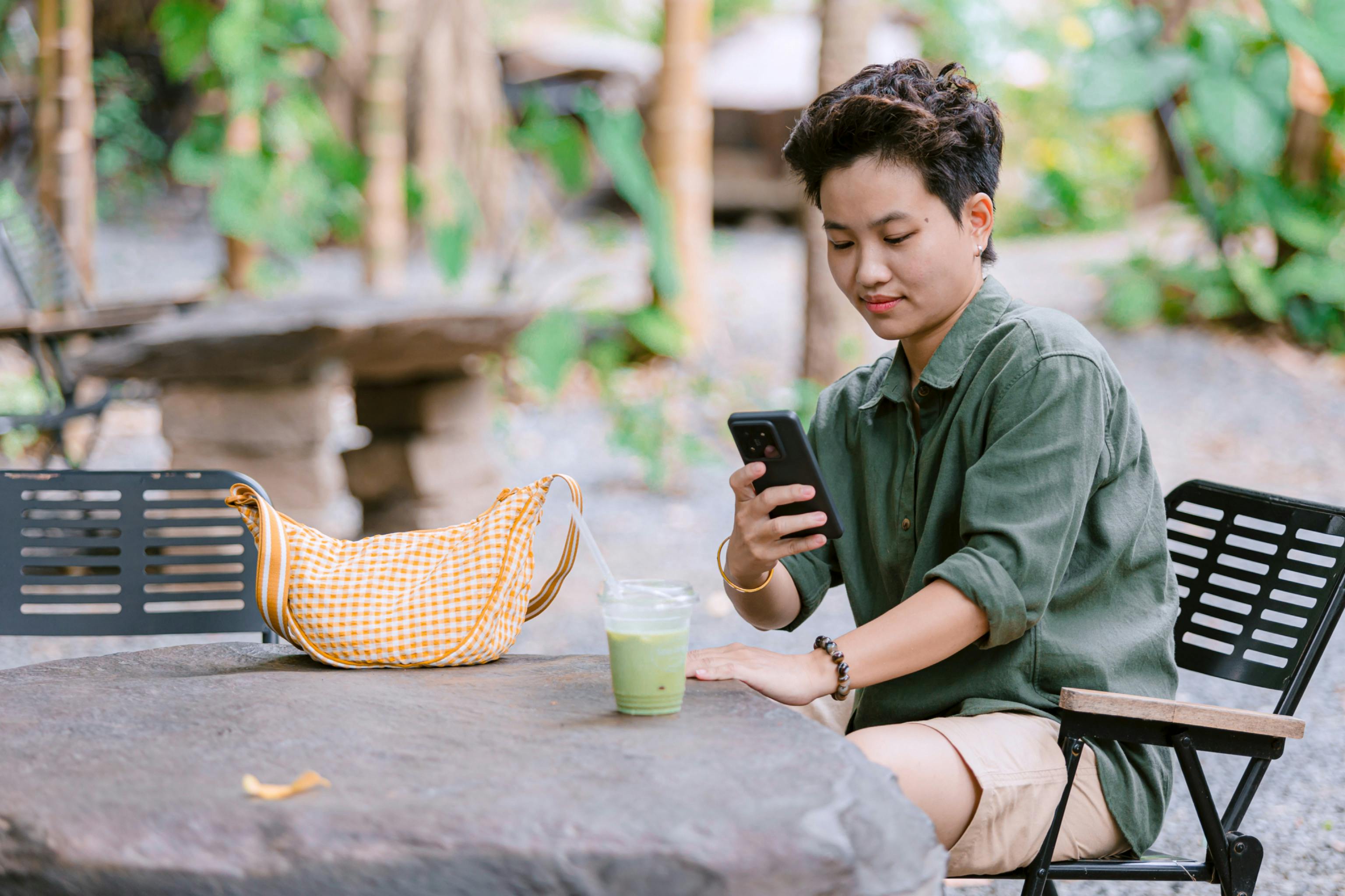 Young Adult Relaxing Outdoors with Phone and Drink · Free Stock Photo