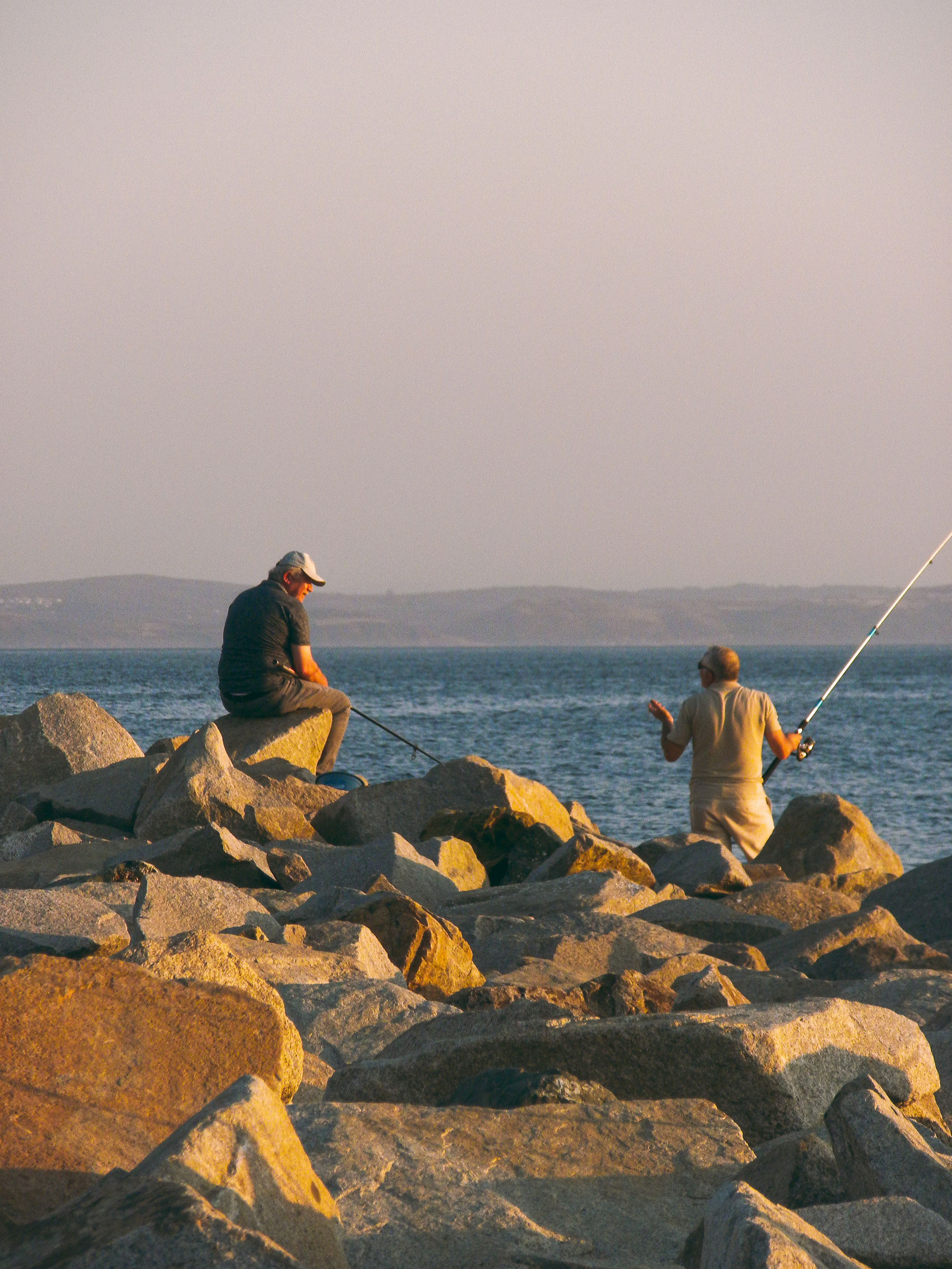 Two Men Fishing on Rocky Shoreline at Sunset · Free Stock Photo