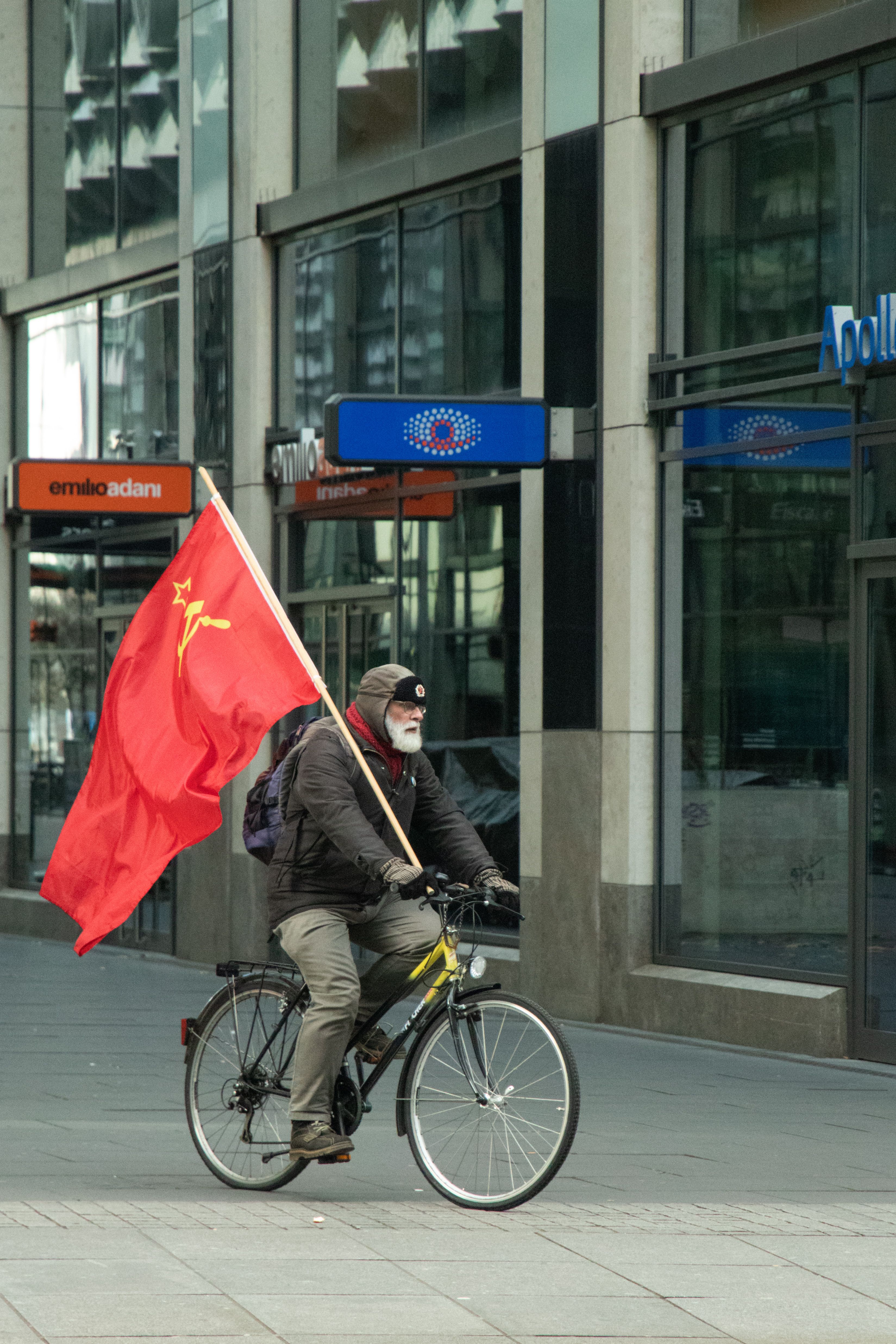 Older Man Riding Bike with Red Flag on City Street · Free Stock Photo