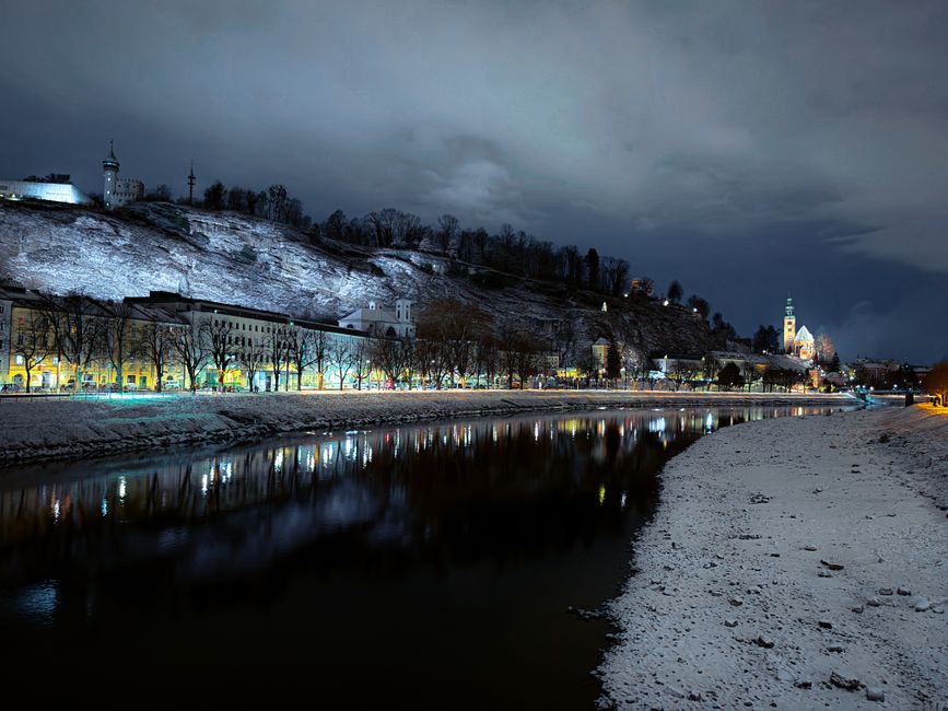 A scenic nighttime view of Salzburg with snow and city lights reflecting on the river.