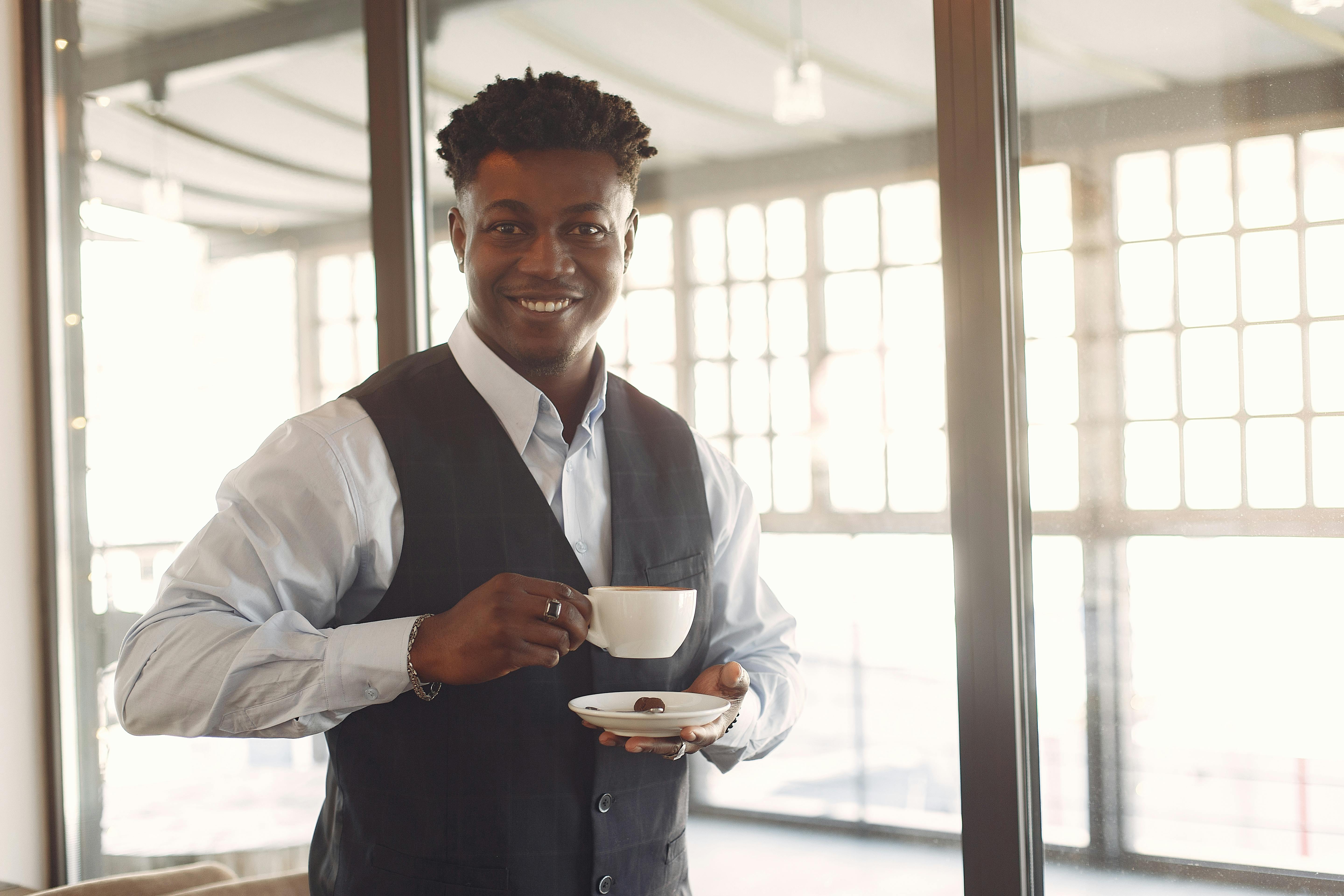 Smiling young ethnic manager drinking coffee while standing in cafe ...