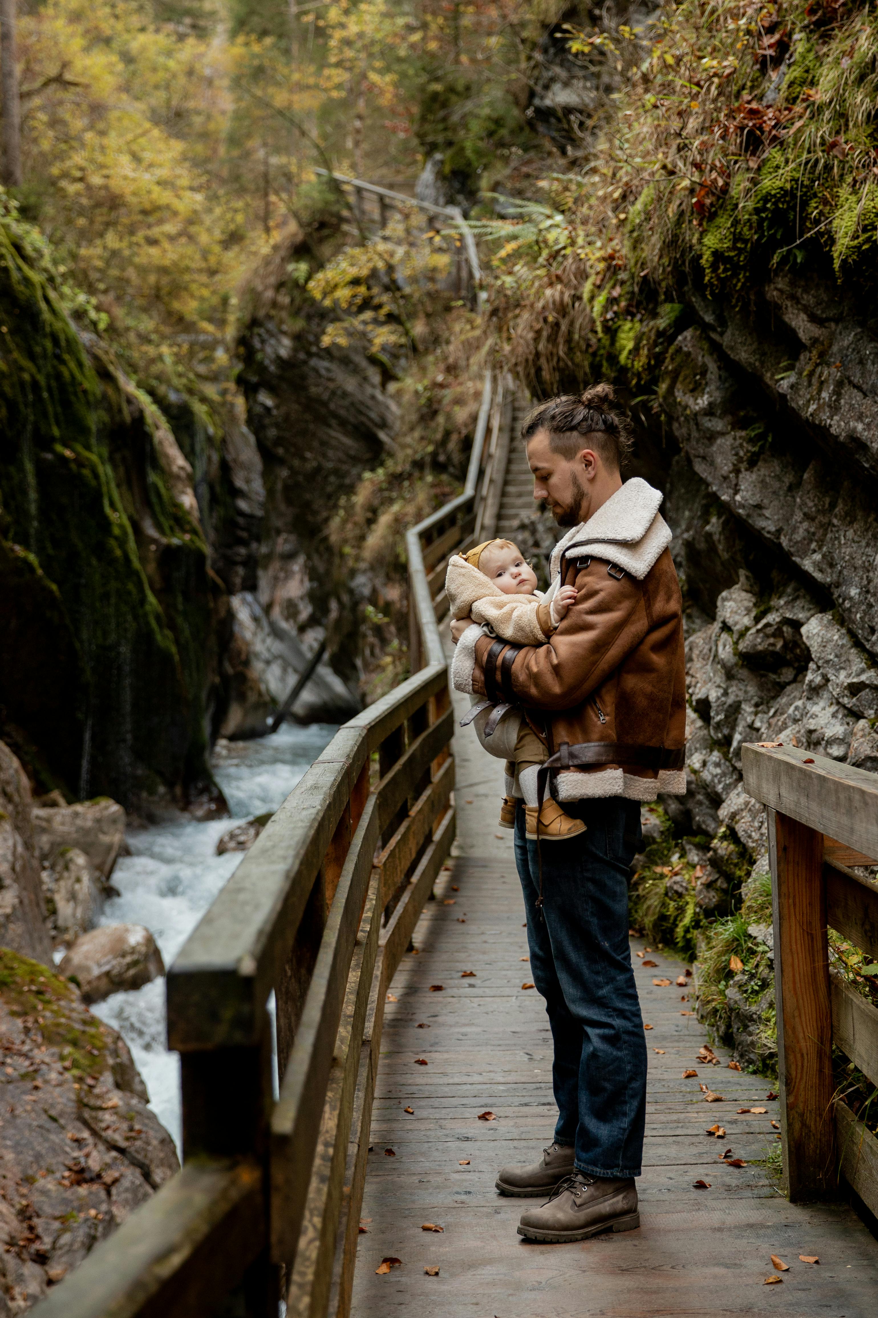 Young father with baby on hand standing on wooden pathway near mountain ...