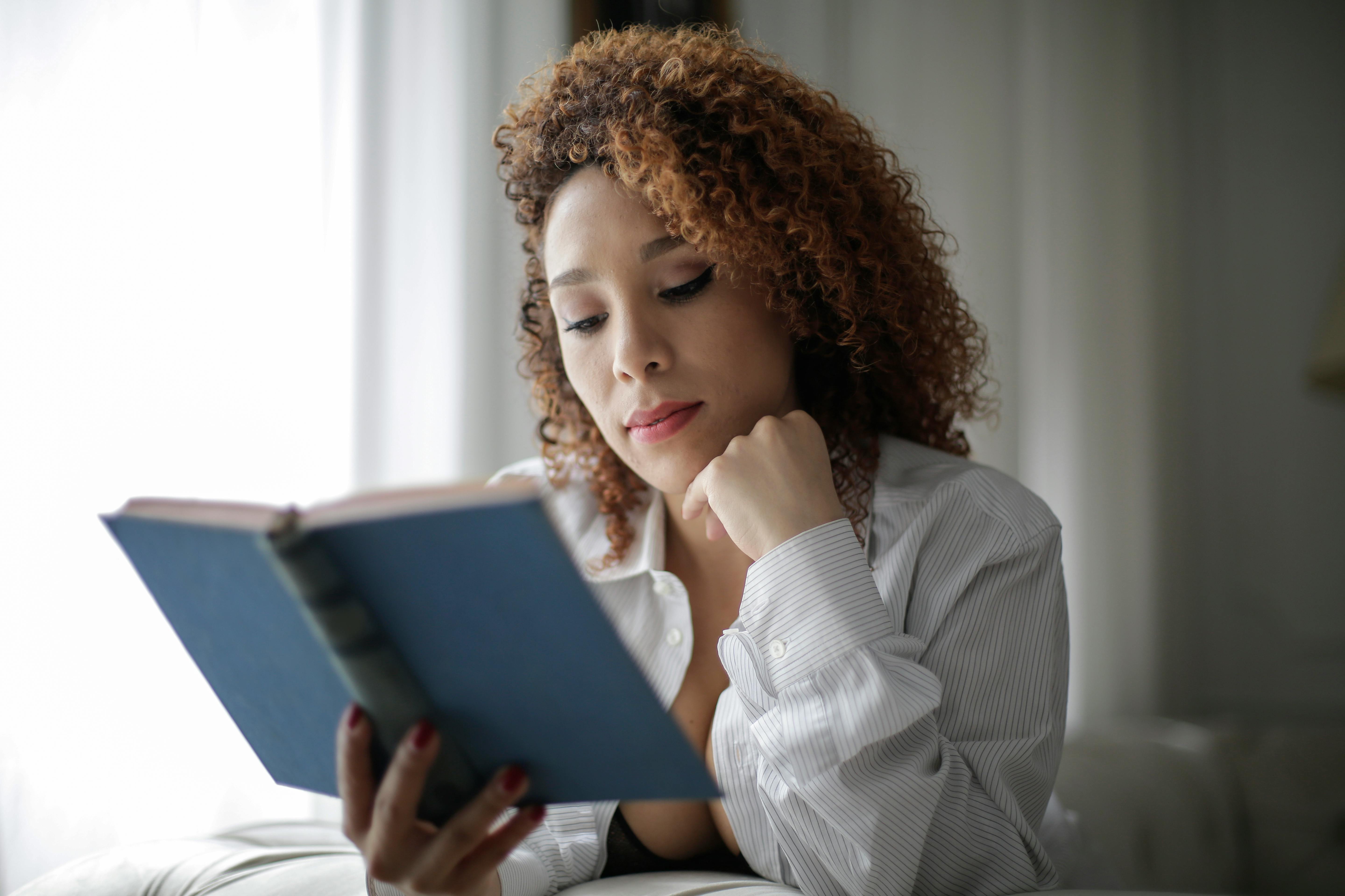 Thoughtful young ethnic lady reading book while resting on sofa at home ...