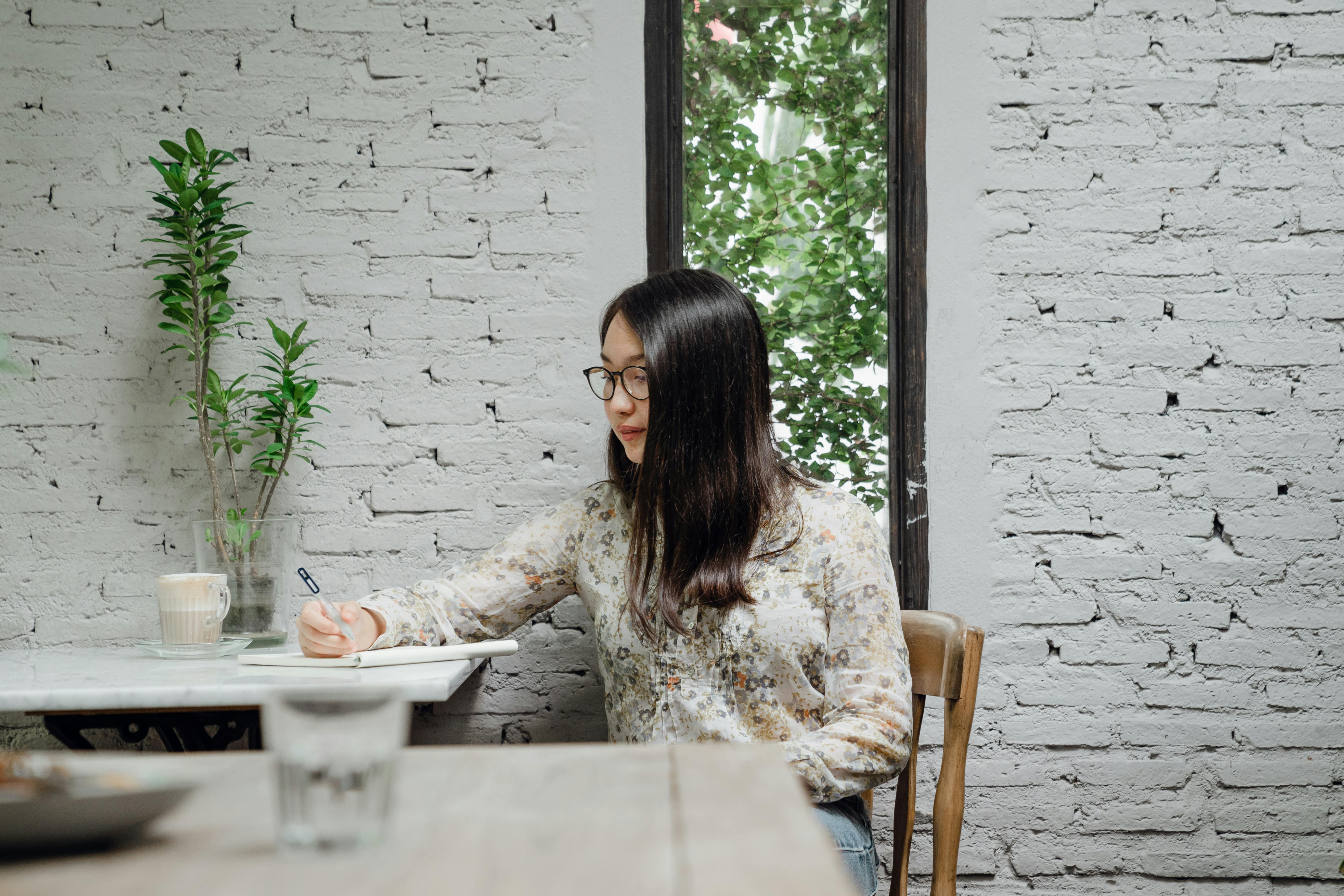Young Asian woman writing in notebook sitting against white brick wall ...