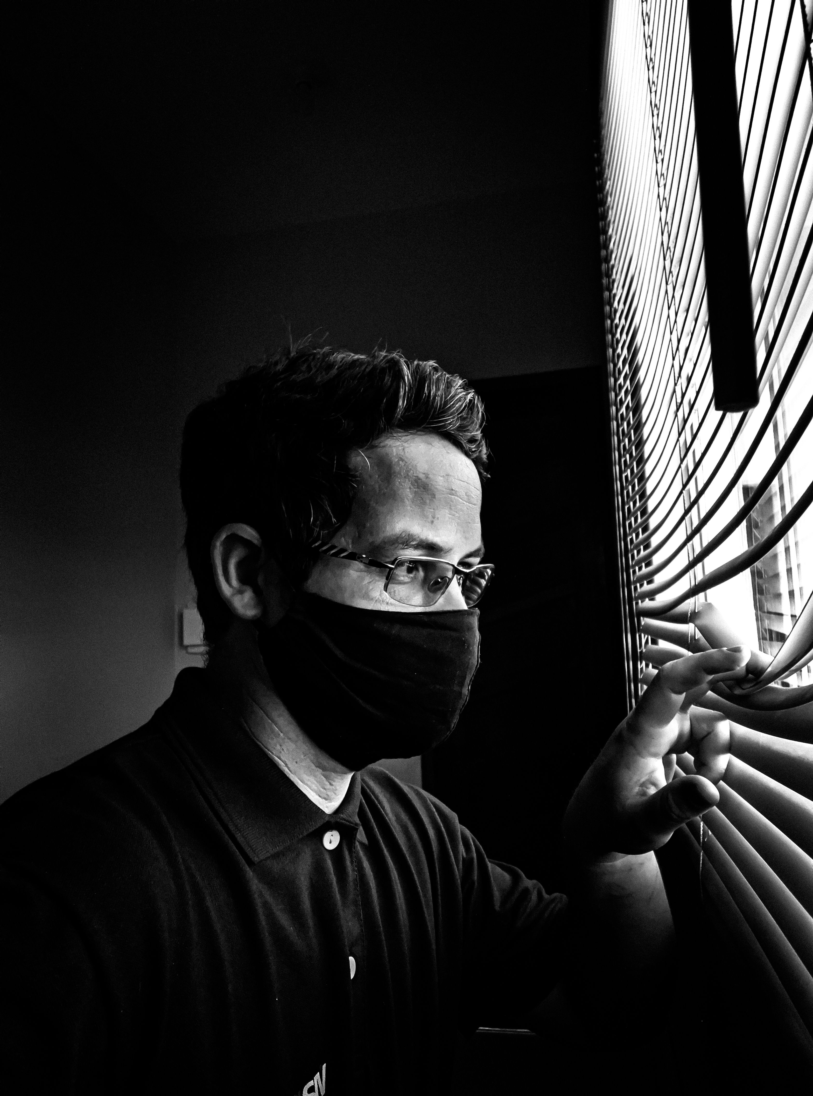 Anonymous man with mask and eyeglasses looking through jalousie in room ...