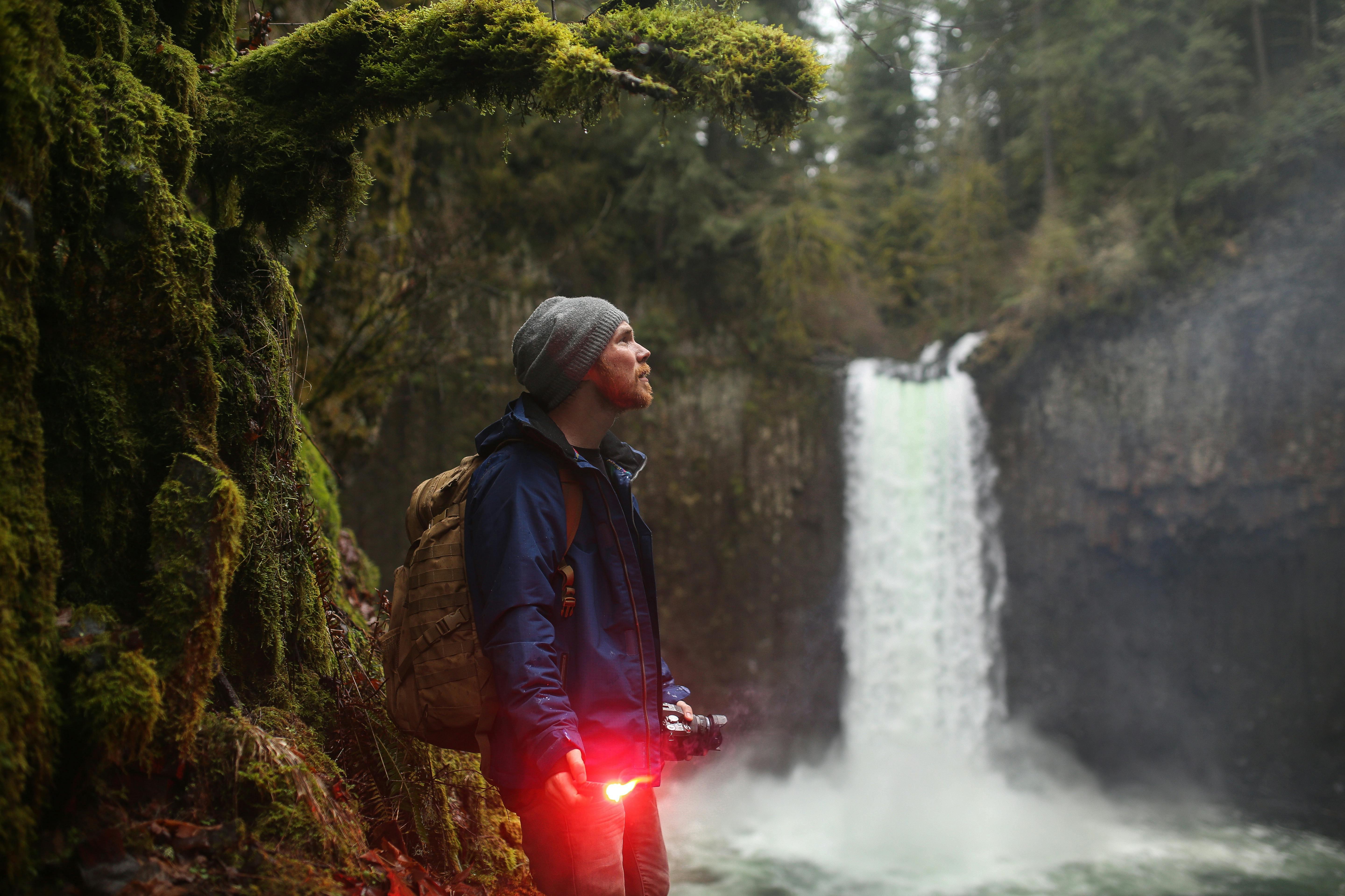 A Man Wearing Blue Jacket Standing Near the Waterfall While Holding a ...