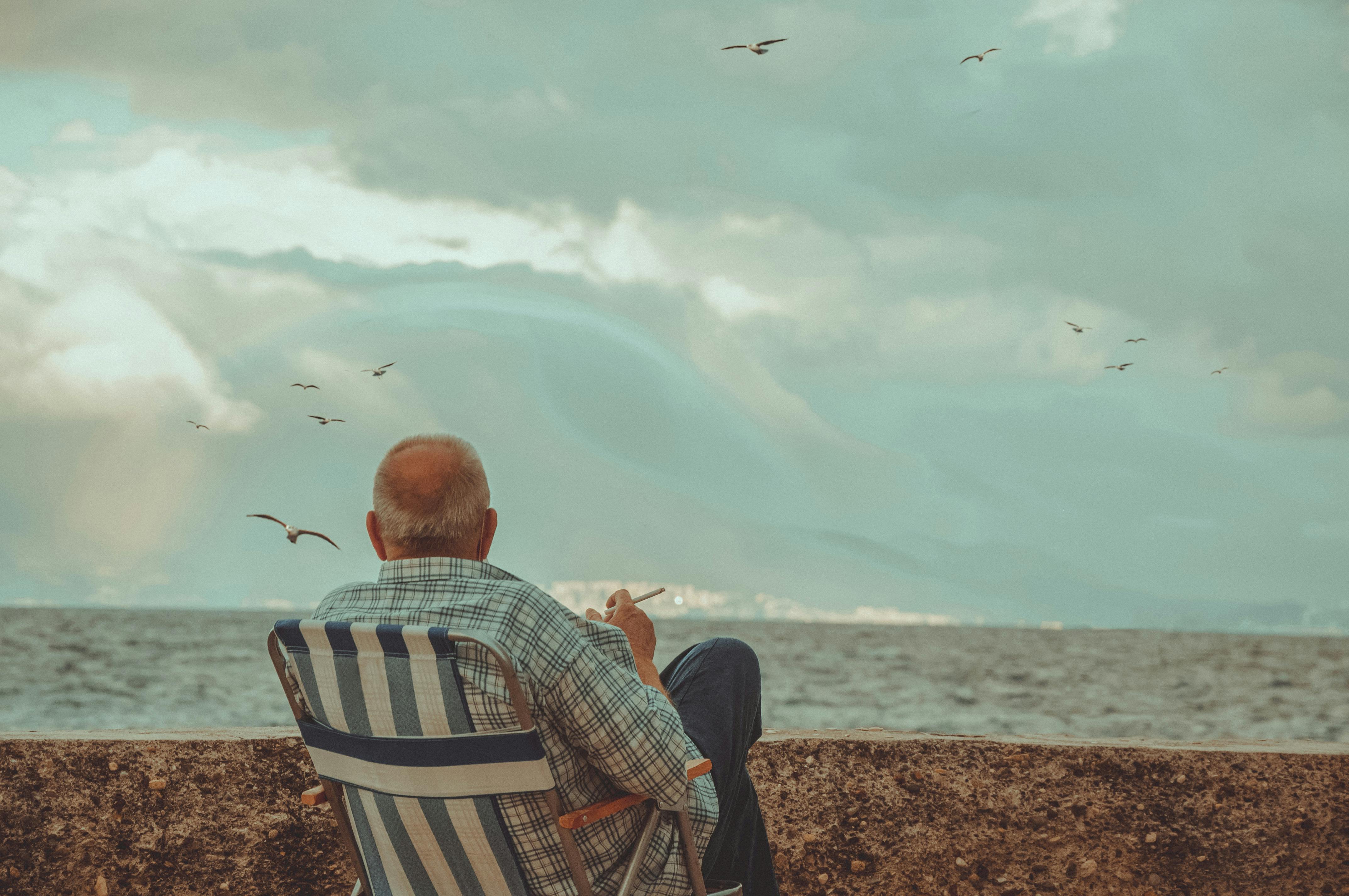 Man Sitting on Chair Looking at Birds Flying Over a Body of Water ...