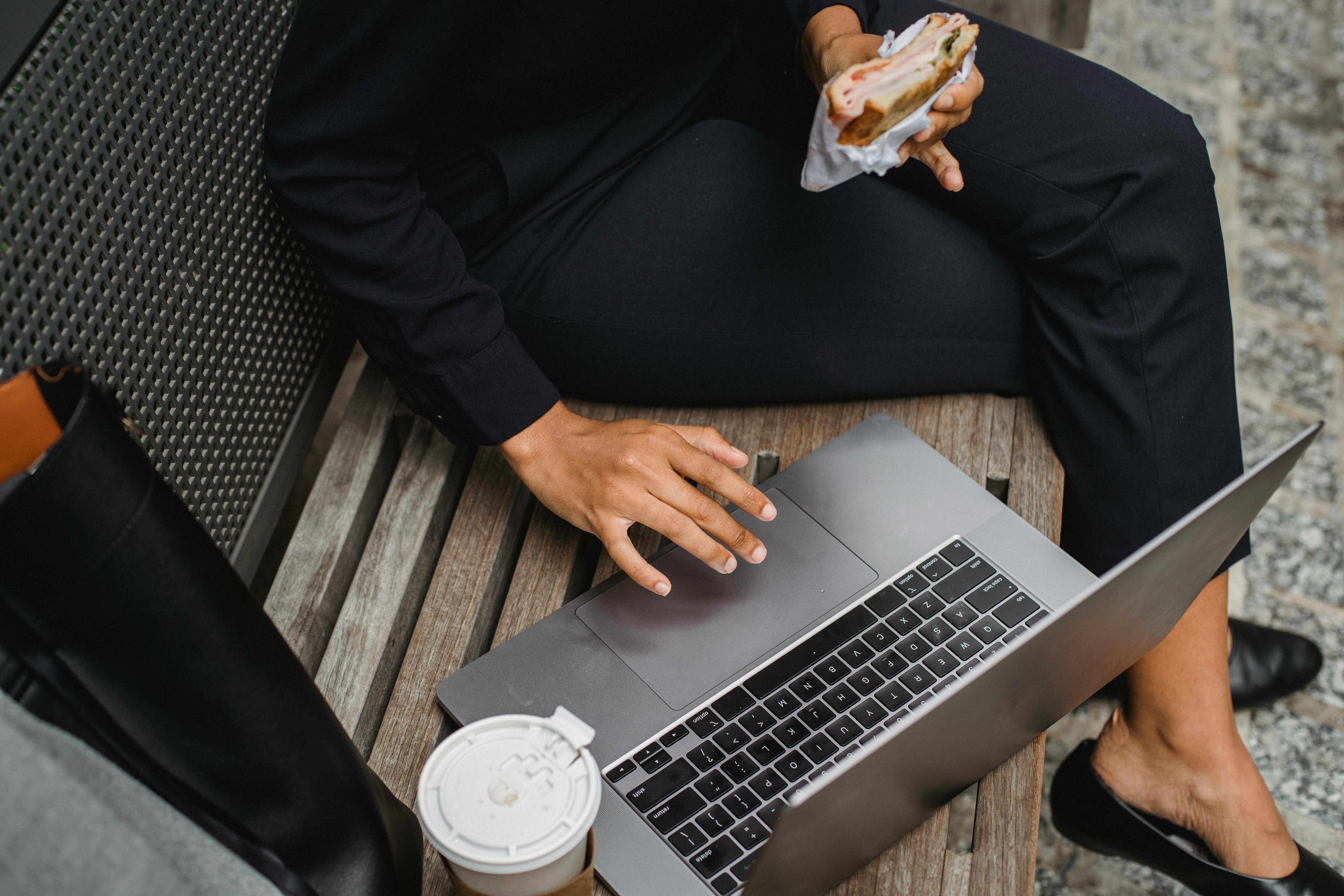A Person Eating While Using a Laptop on the Wooden Bench · Free Stock Photo
