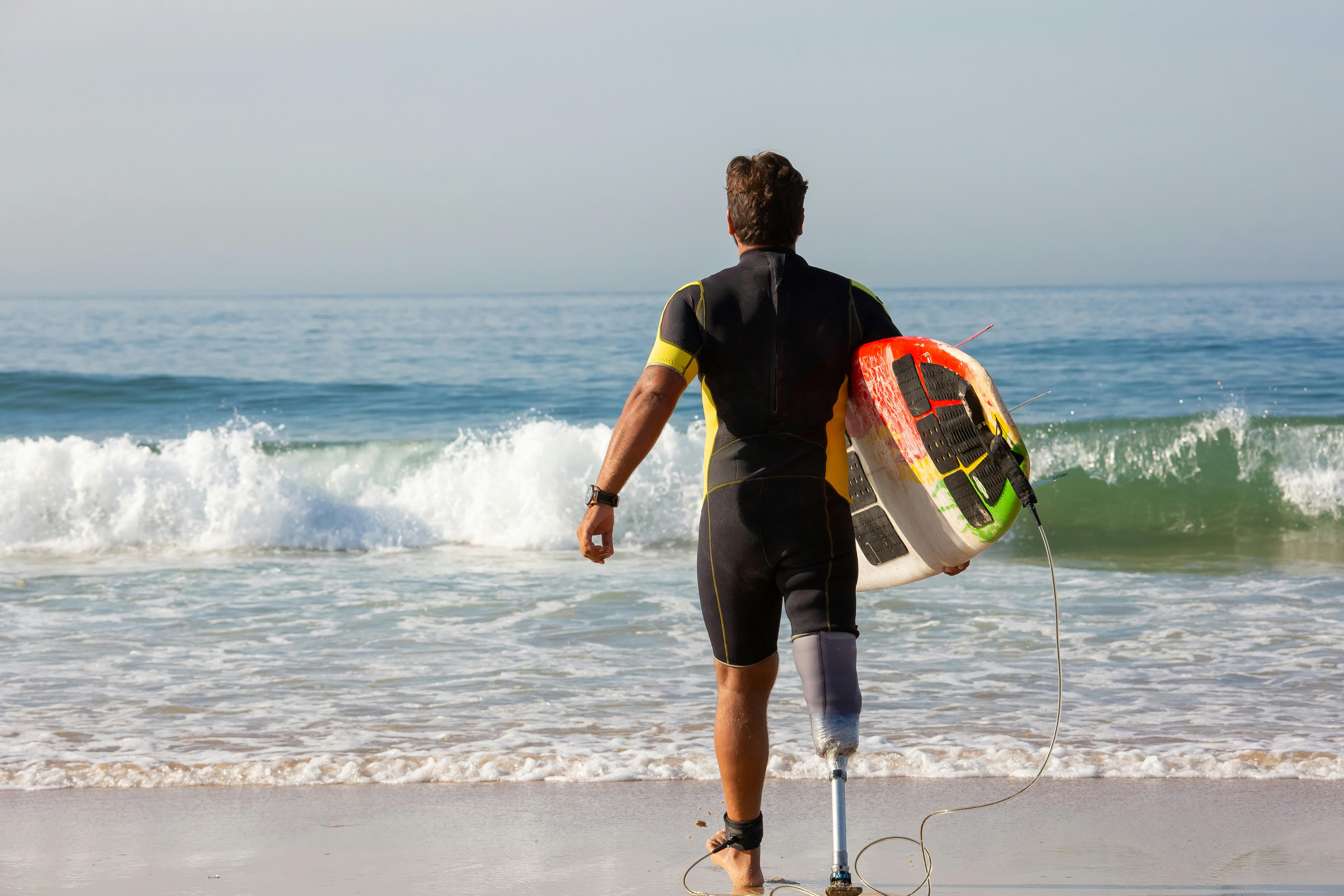 Unrecognizable male surfer amputee with surfboard walking towards sea ...
