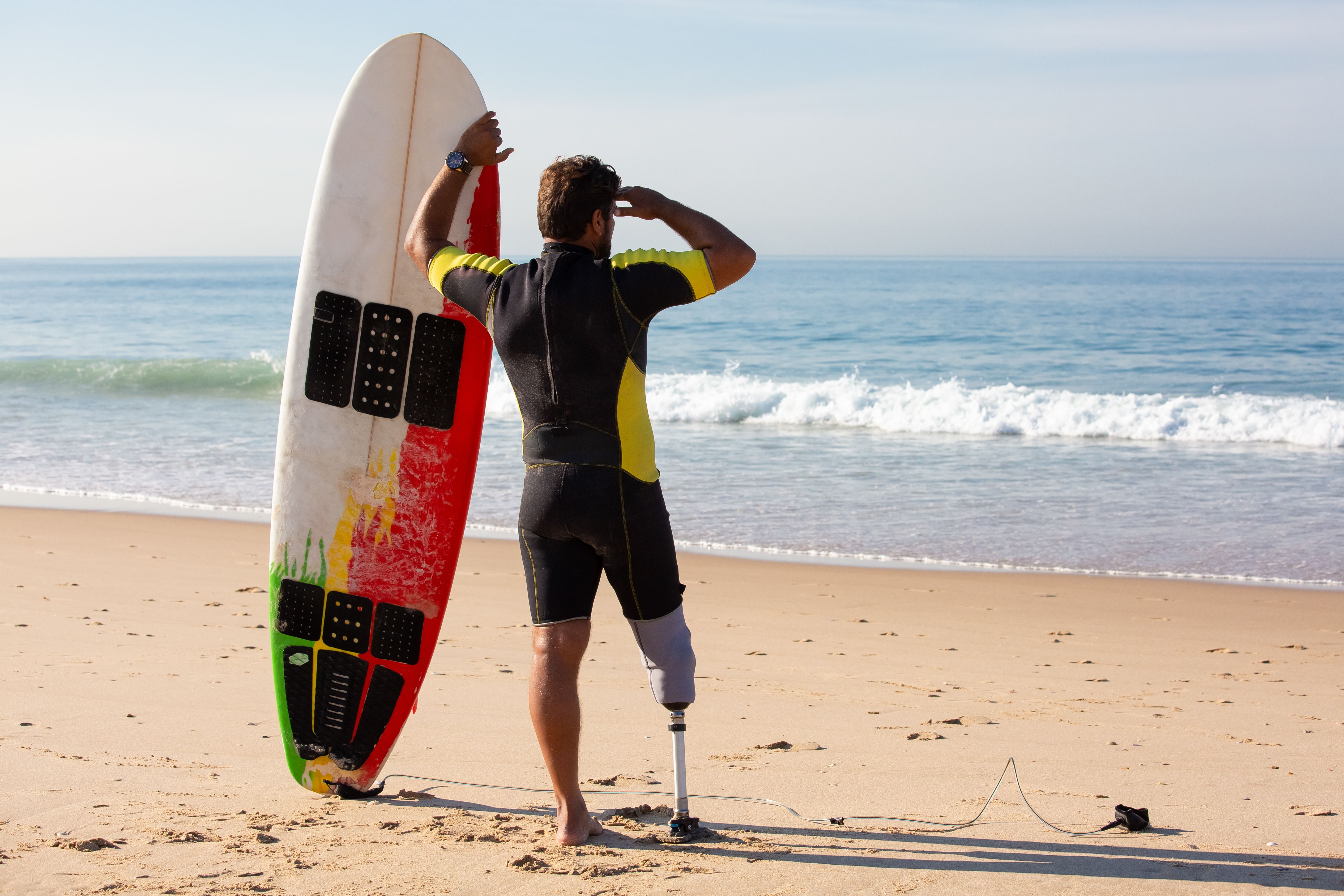 Unrecognizable male amputee surfer with surfboard standing on seashore ...