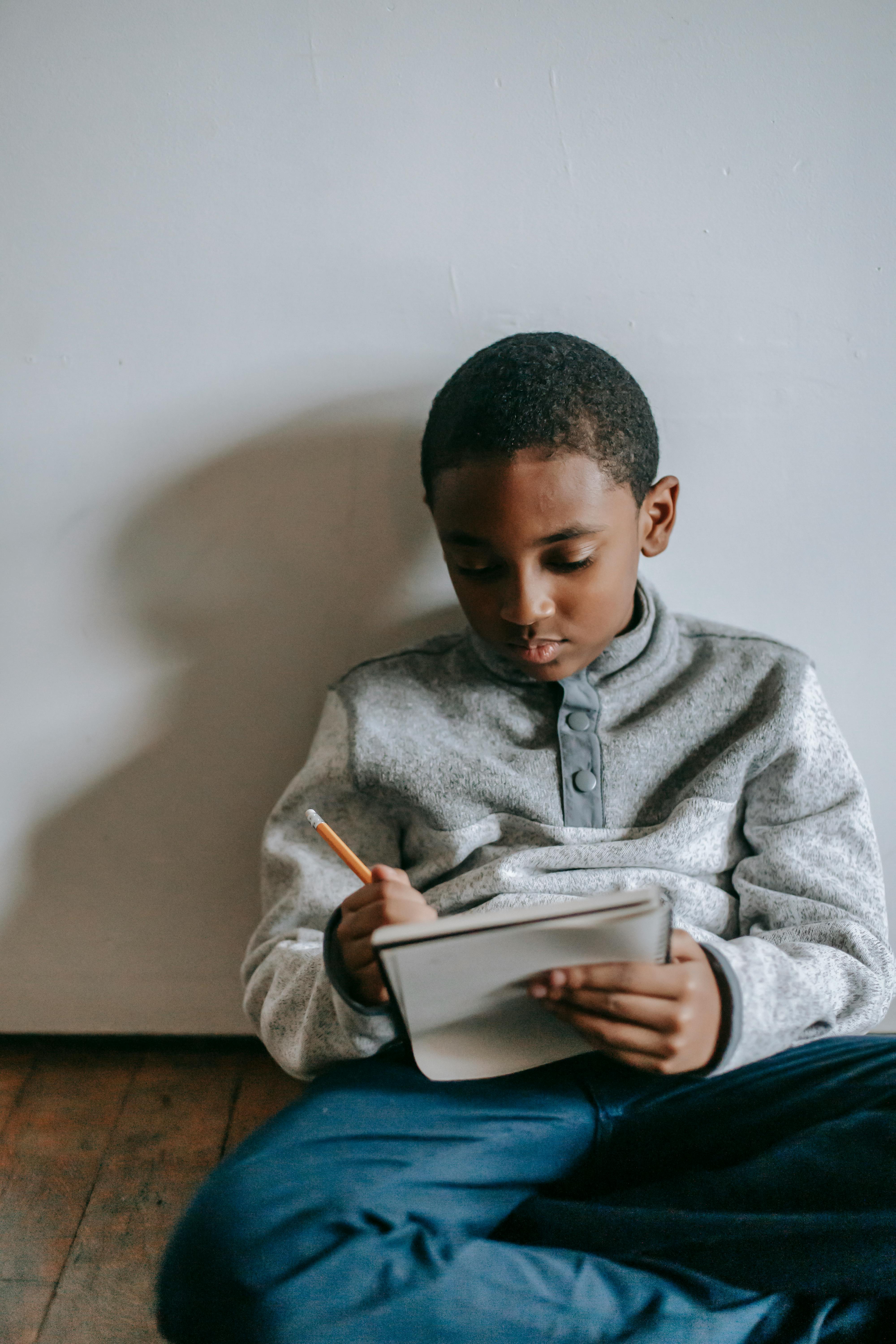 Concentrated black schoolchild taking notes in notebook near wall ...