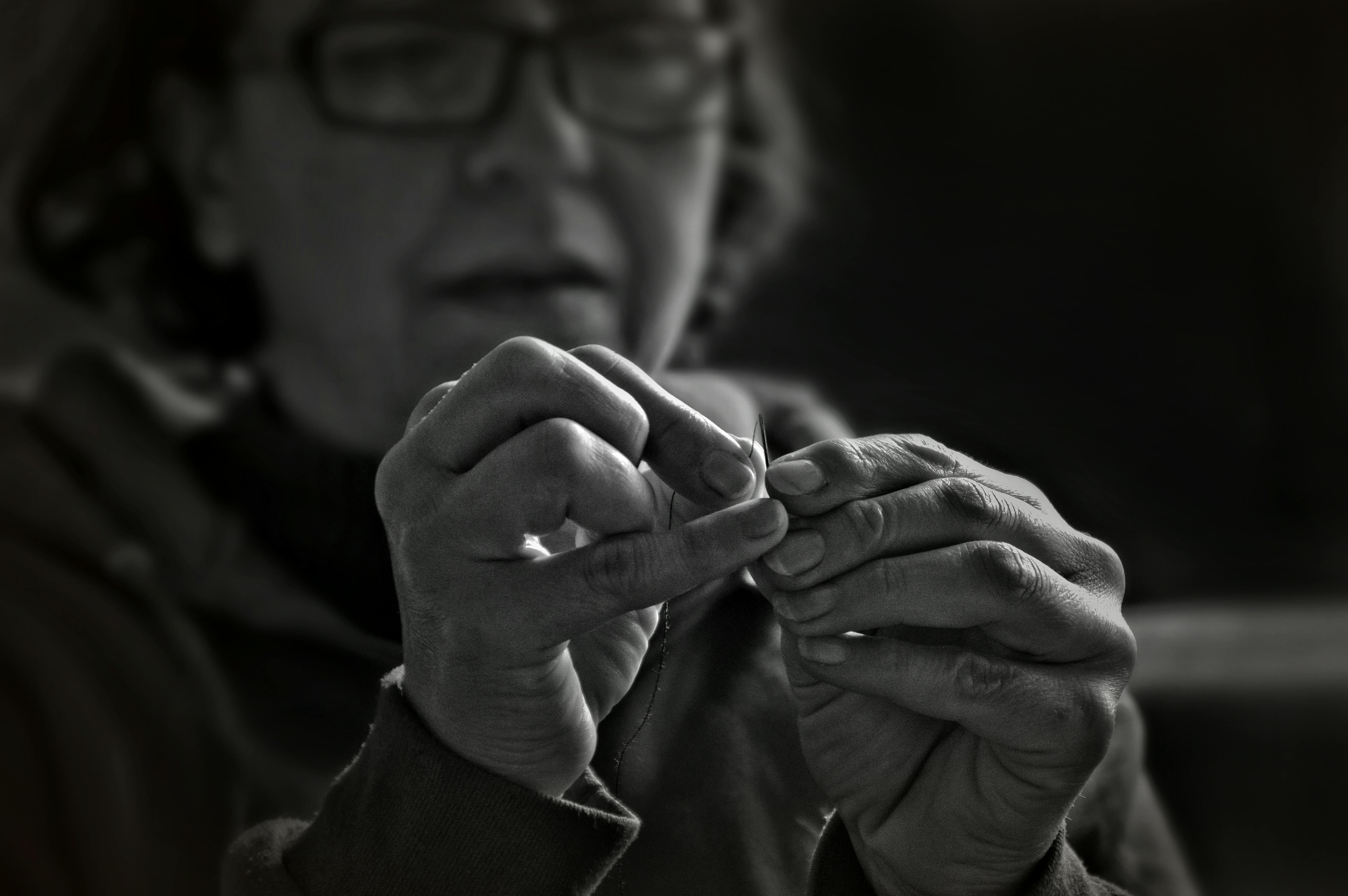 Grayscale Photo of a Woman Wearing Eyeglasses Inserting the Thread on ...