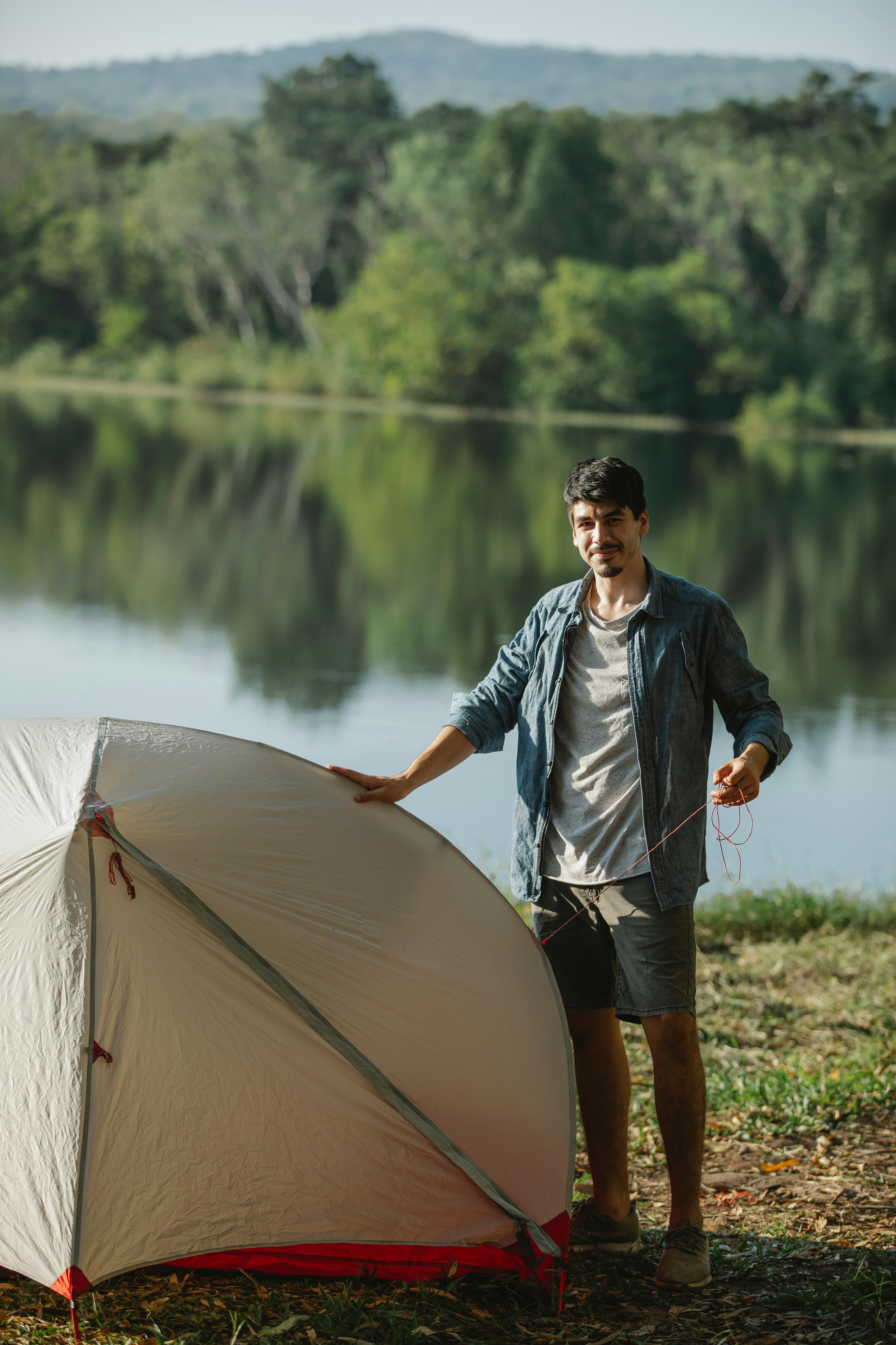 Smiling young guy setting up camping tent on lake shore after trekking ...