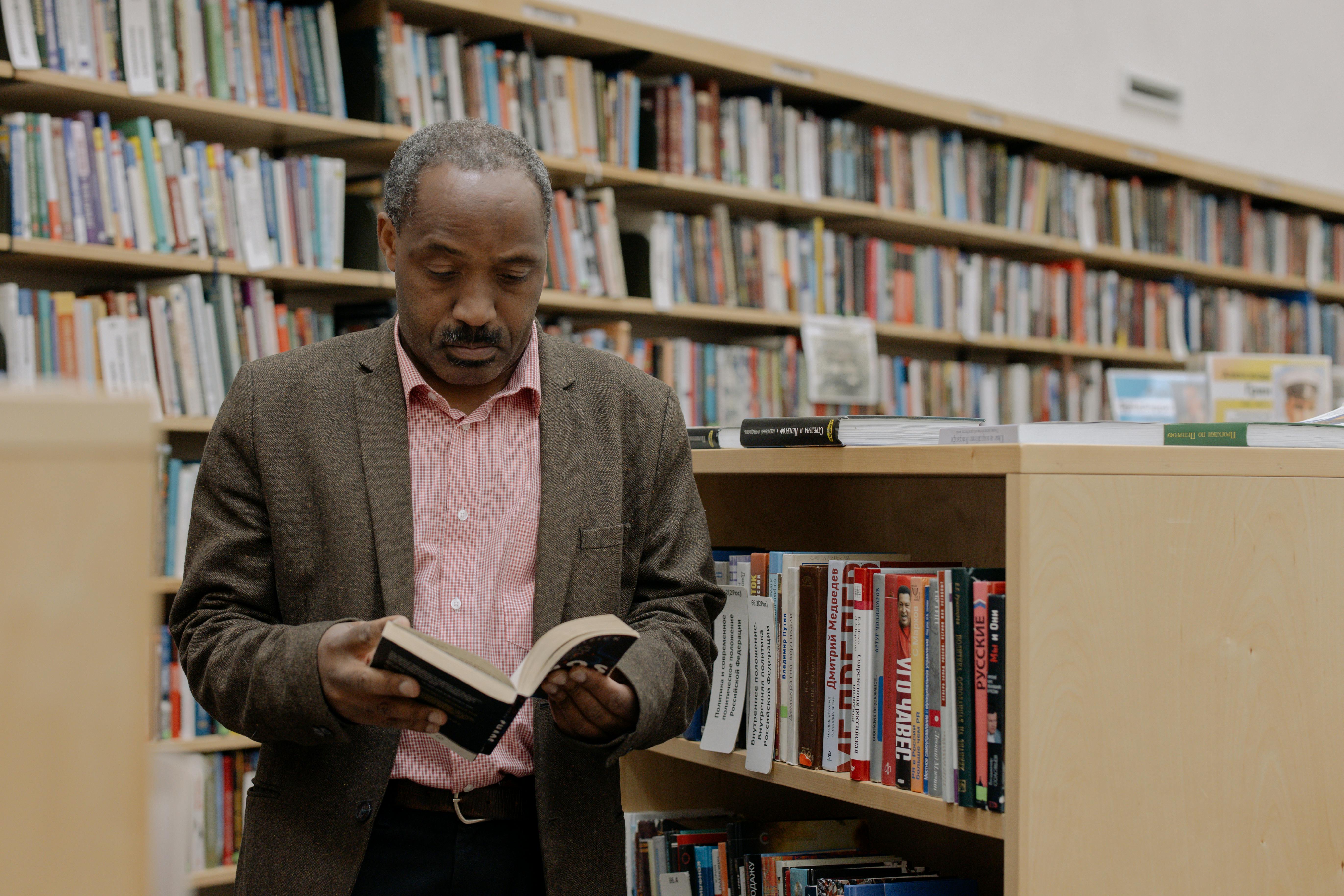 Man Standing by a Bookshelf in a Library and Reading a Book · Free ...