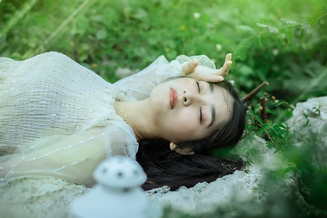 A Woman In White Long Sleeves Lying On Green Grass With Her Eyes Closed