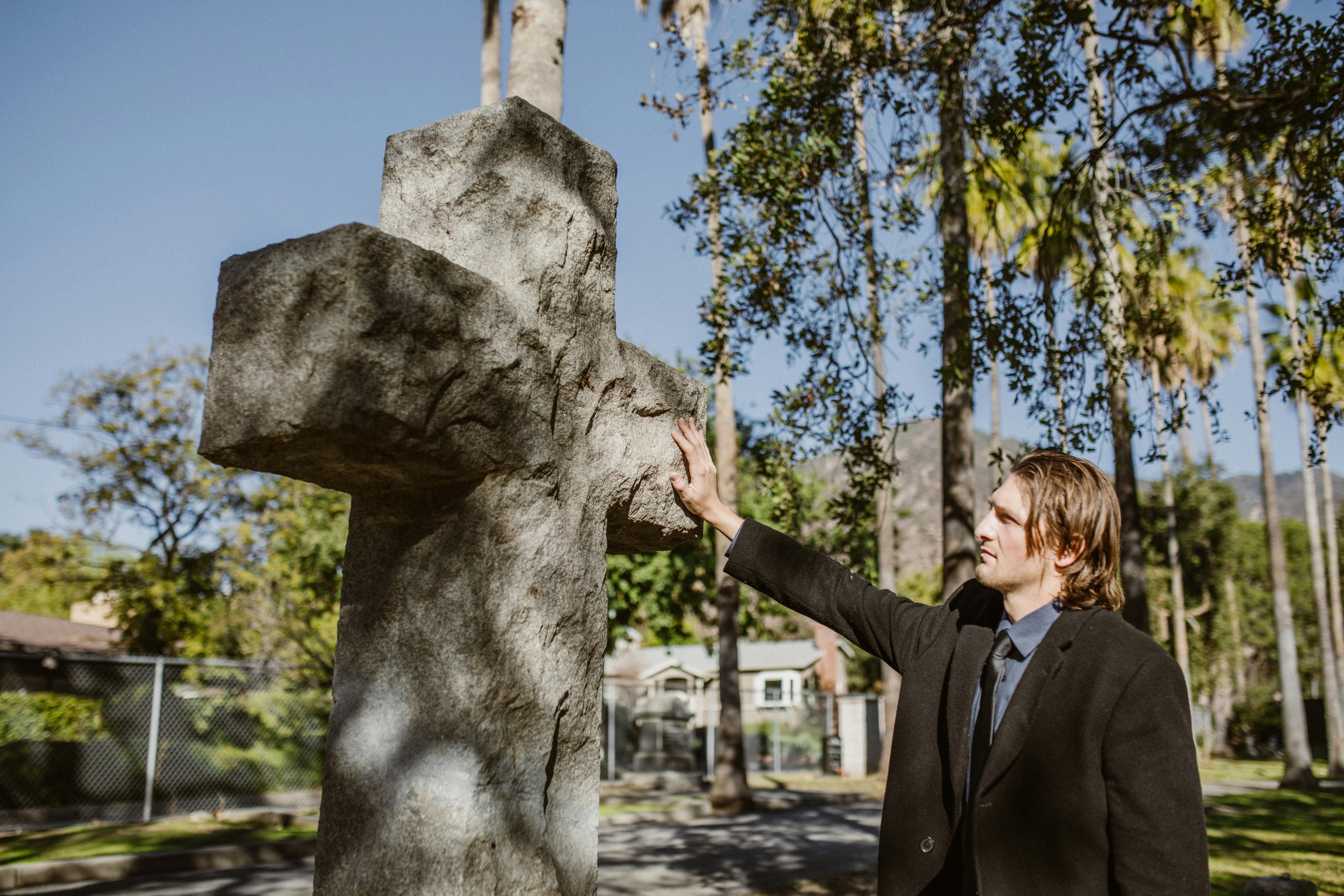 A Man in a Black Coat Touching a Stone Crucifix · Free Stock Photo