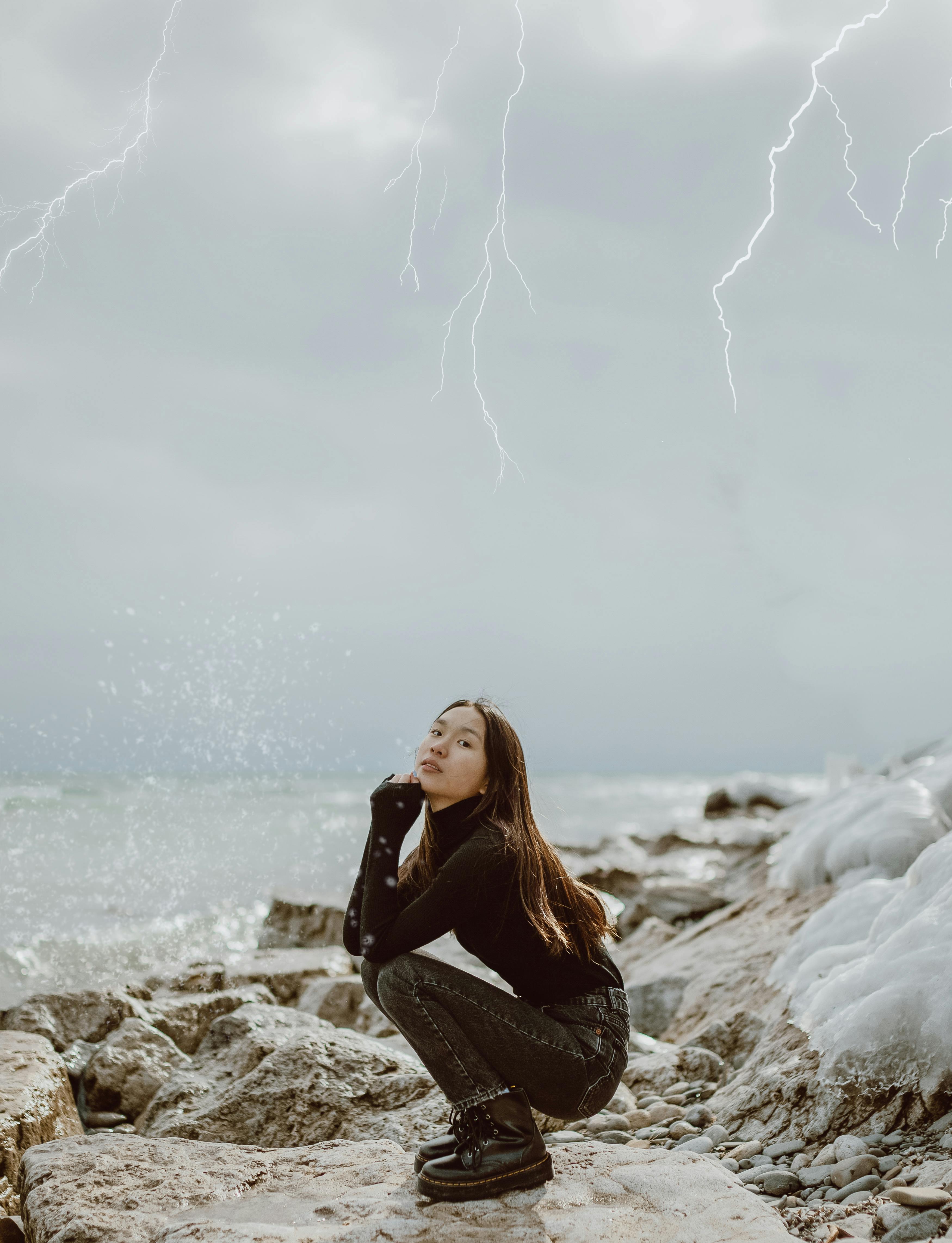 Confident young ethnic woman sitting on haunches on rocky seashore ...