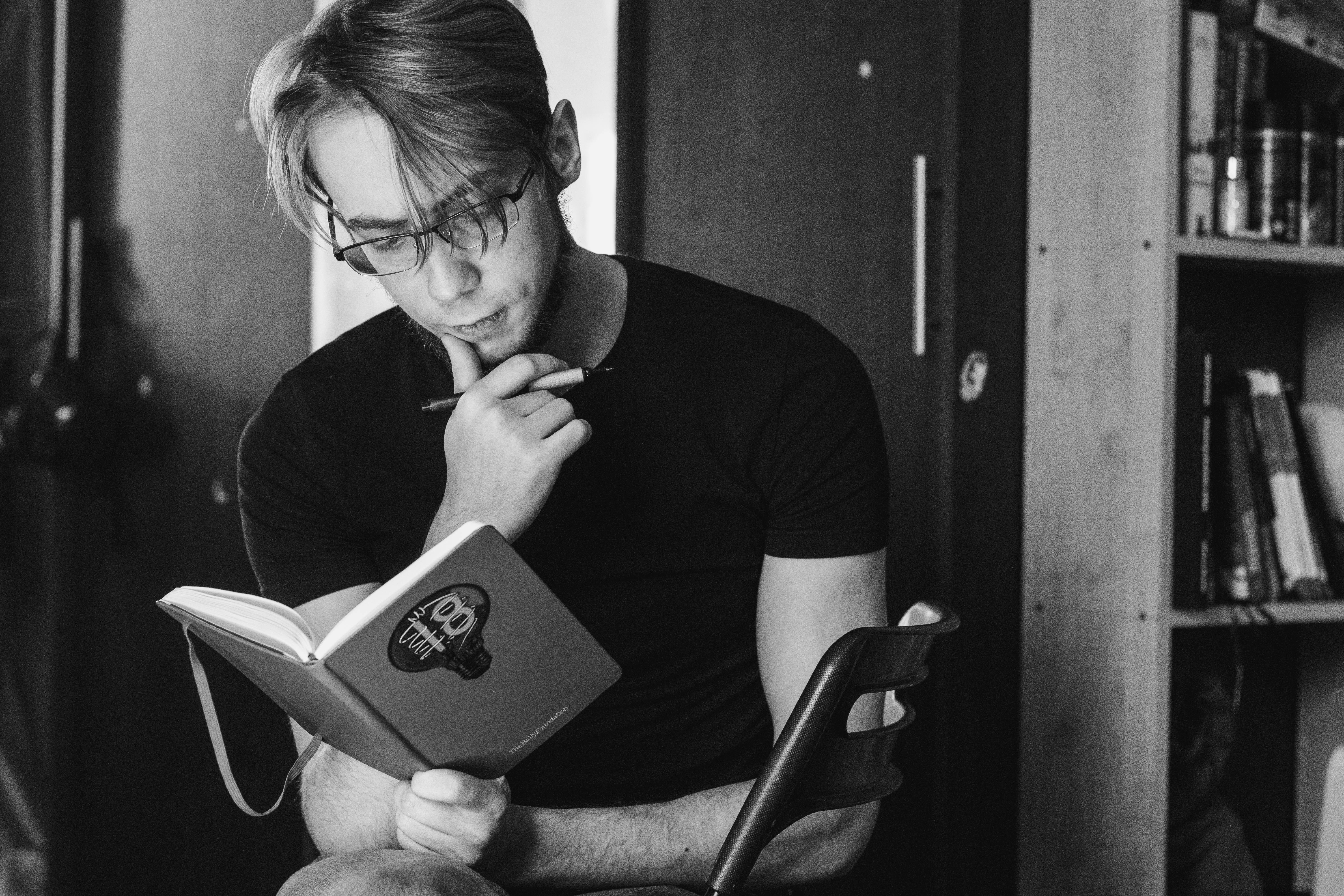 Grayscale Photo of a Man Sitting and Reading a Notebook · Free Stock Photo