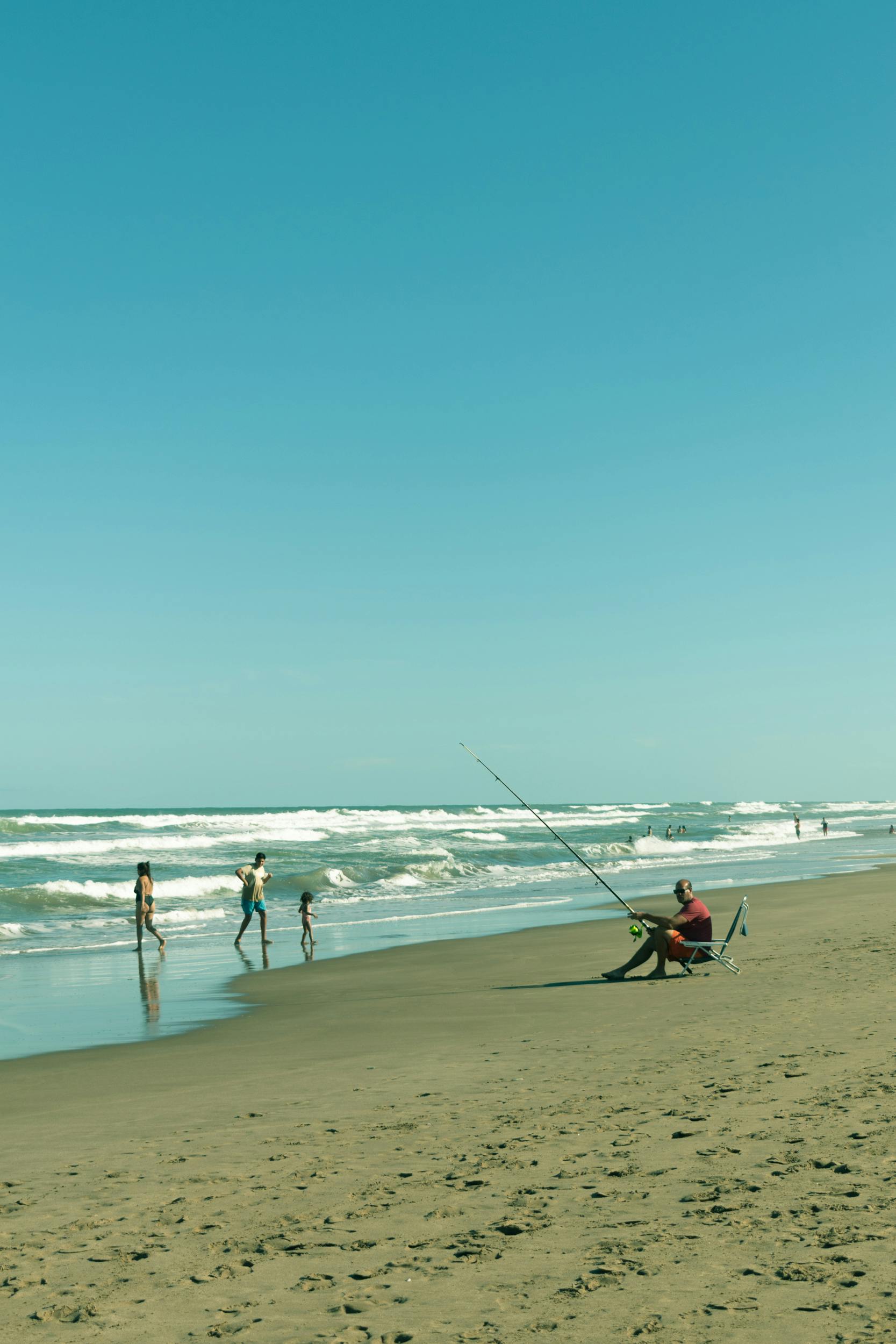 People Walking on Beach Shore Near a Man Fishing · Free Stock Photo