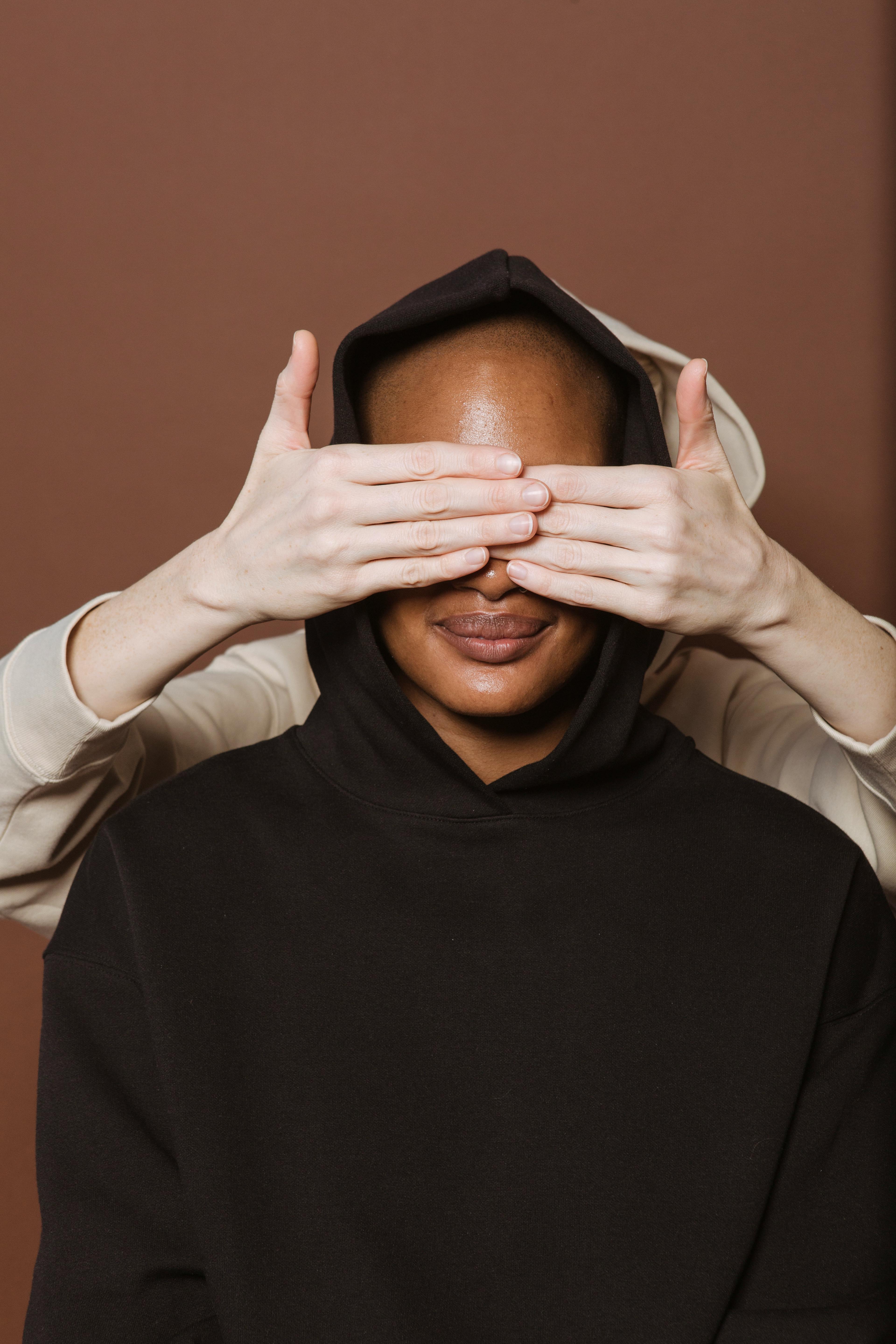 Anonymous black woman covering eyes of female friend in brown studio ...