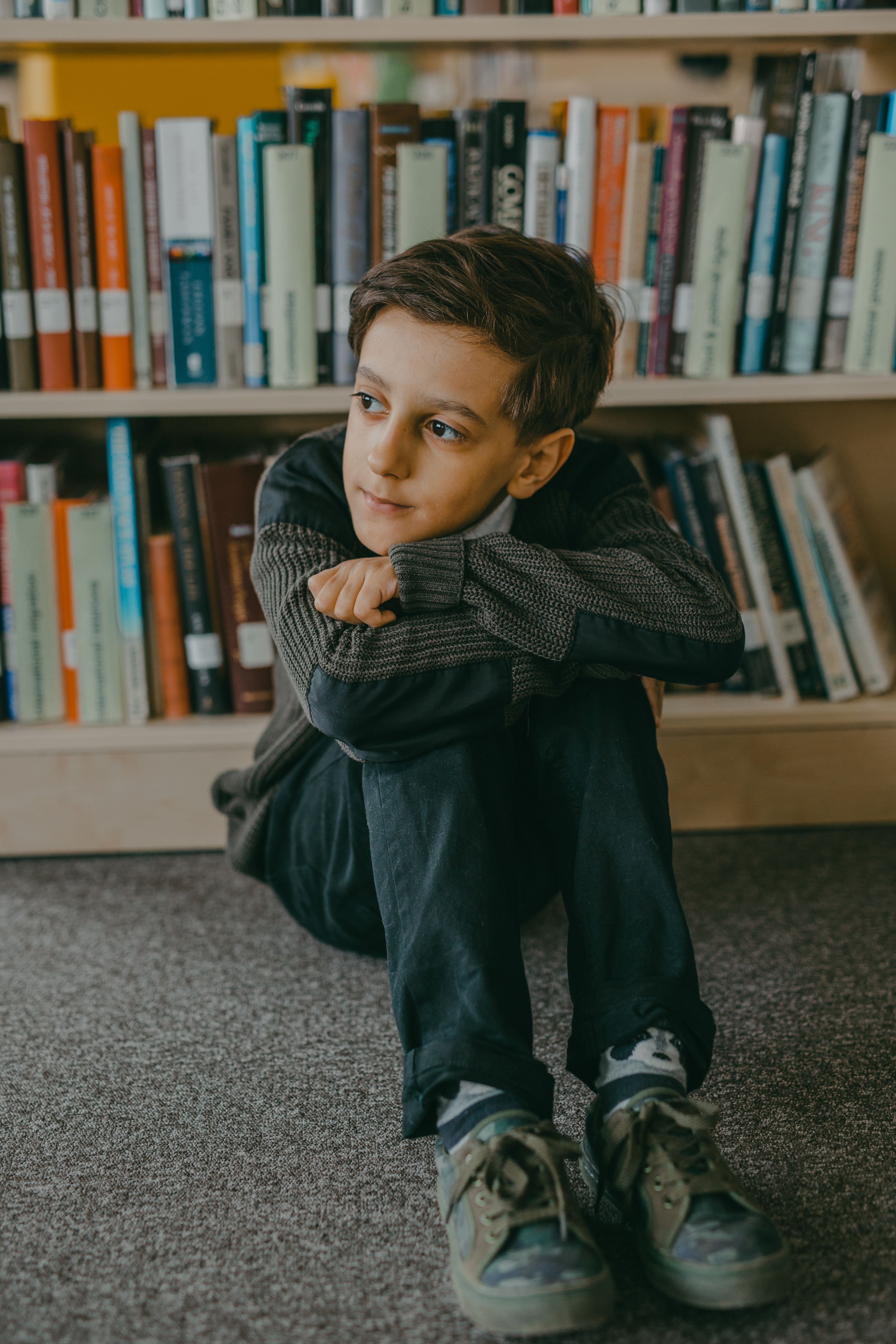 A Boy Wearing Sweater and Pants Sitting on the Floor Near Bookcase ...