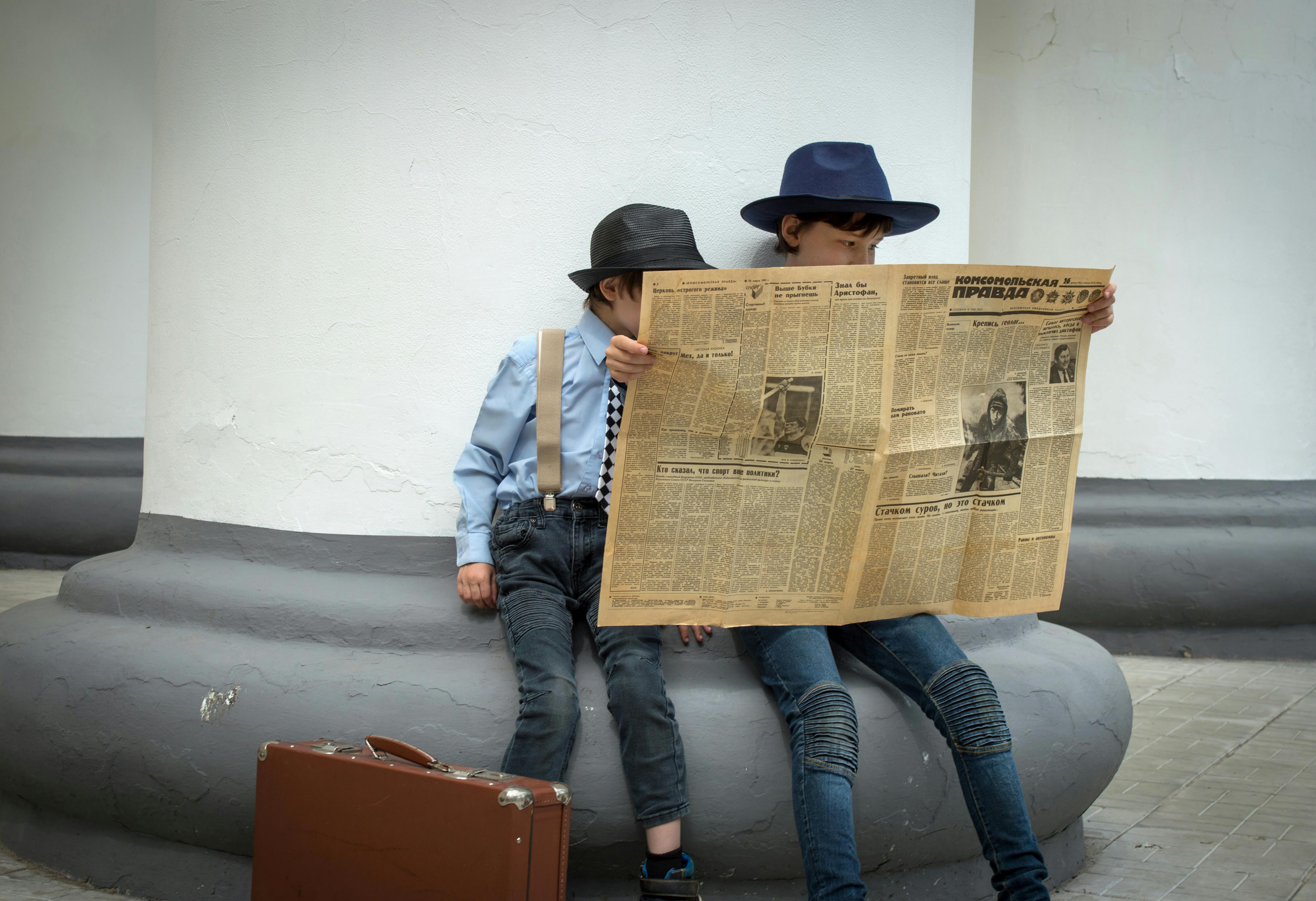 Children Reading Newspaper while Sitting near a Wall · Free Stock Photo