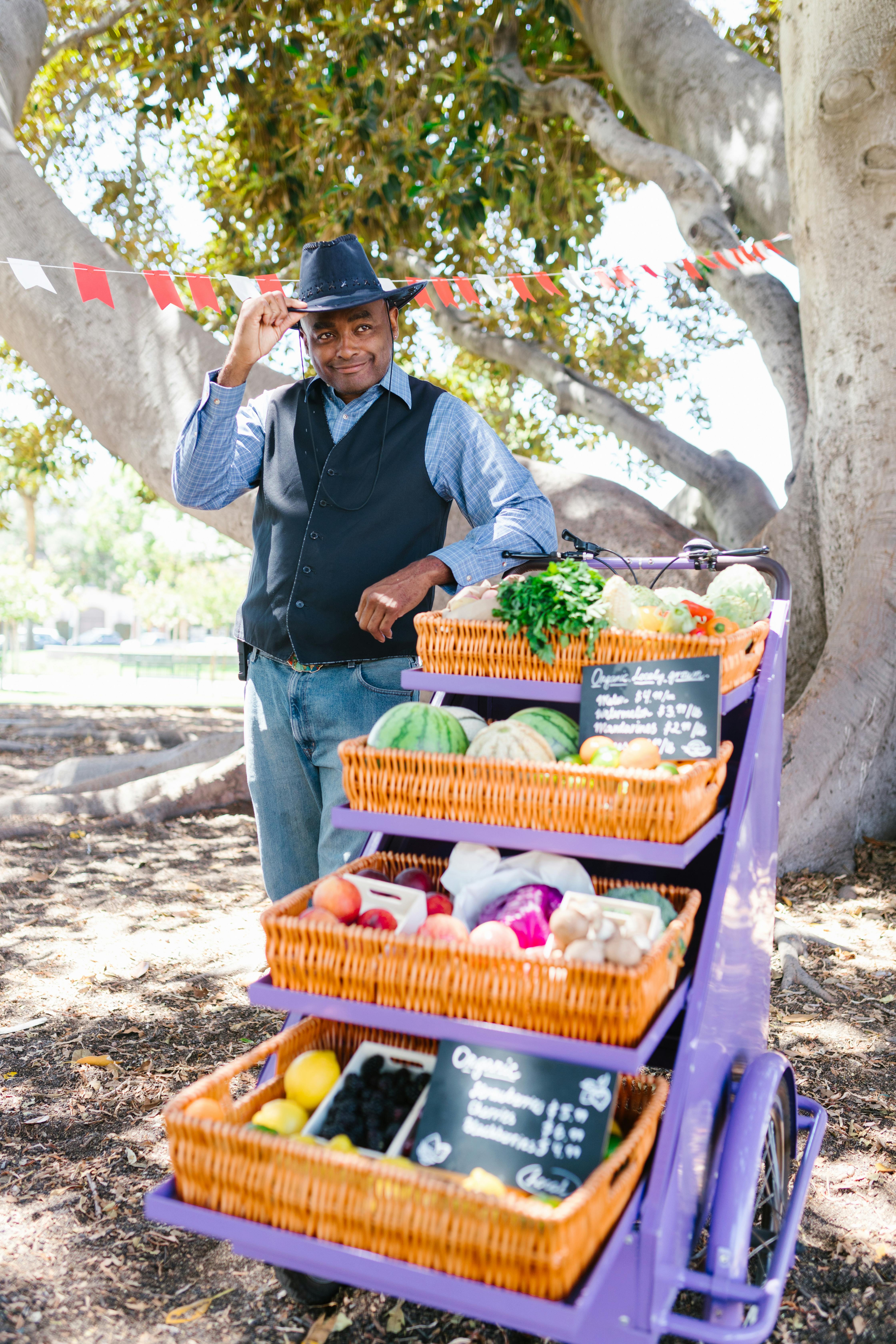 Man Wearing a Vest and Hat Standing Near a Fruit Stand · Free Stock Photo