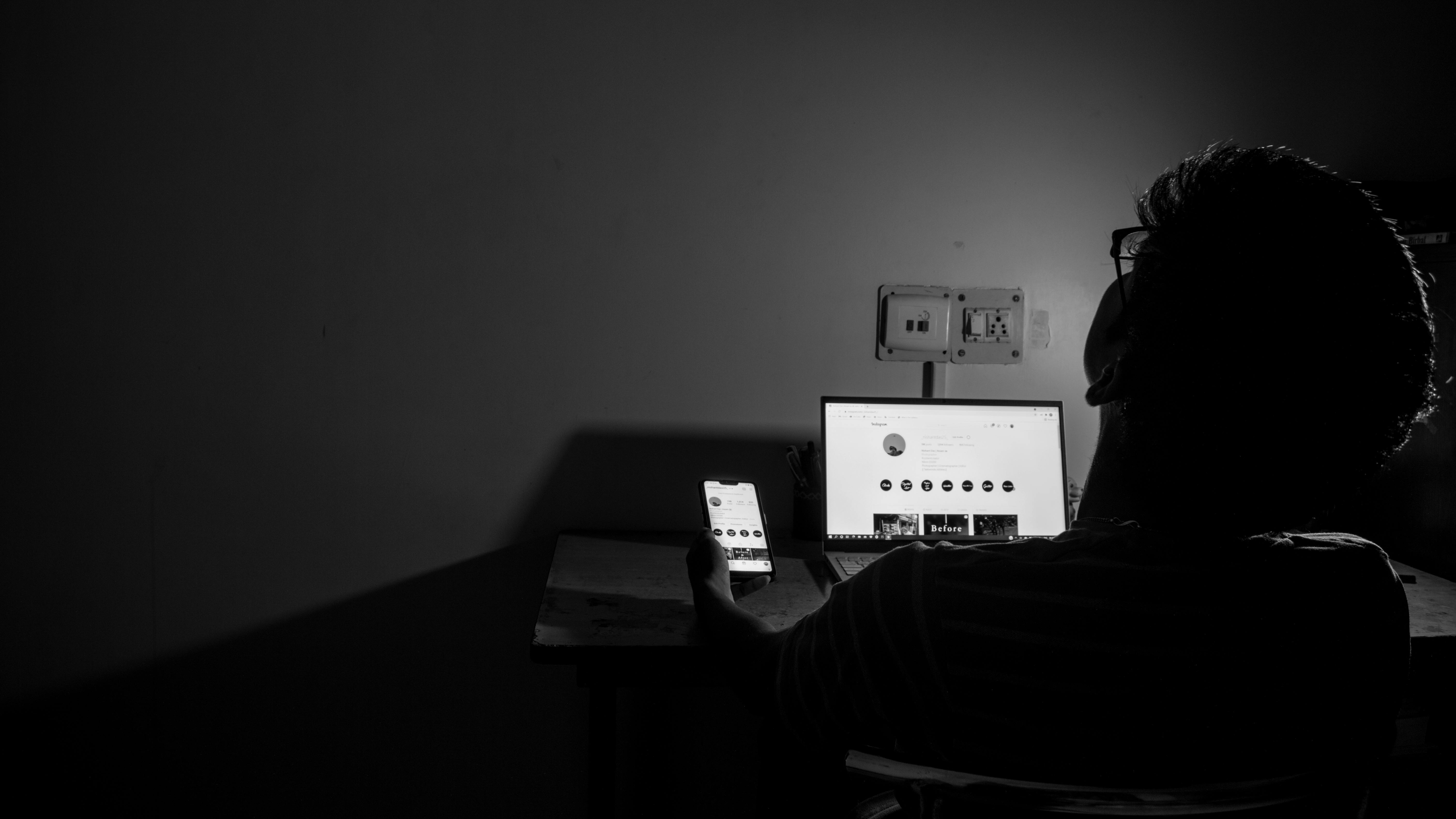 A Grayscale of a Man Using His Smartphone and a His Laptop in a Dark ...