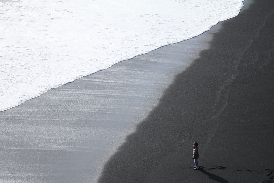 A child stands alone on a striking black sand beach in Iceland, facing the ocean waves.