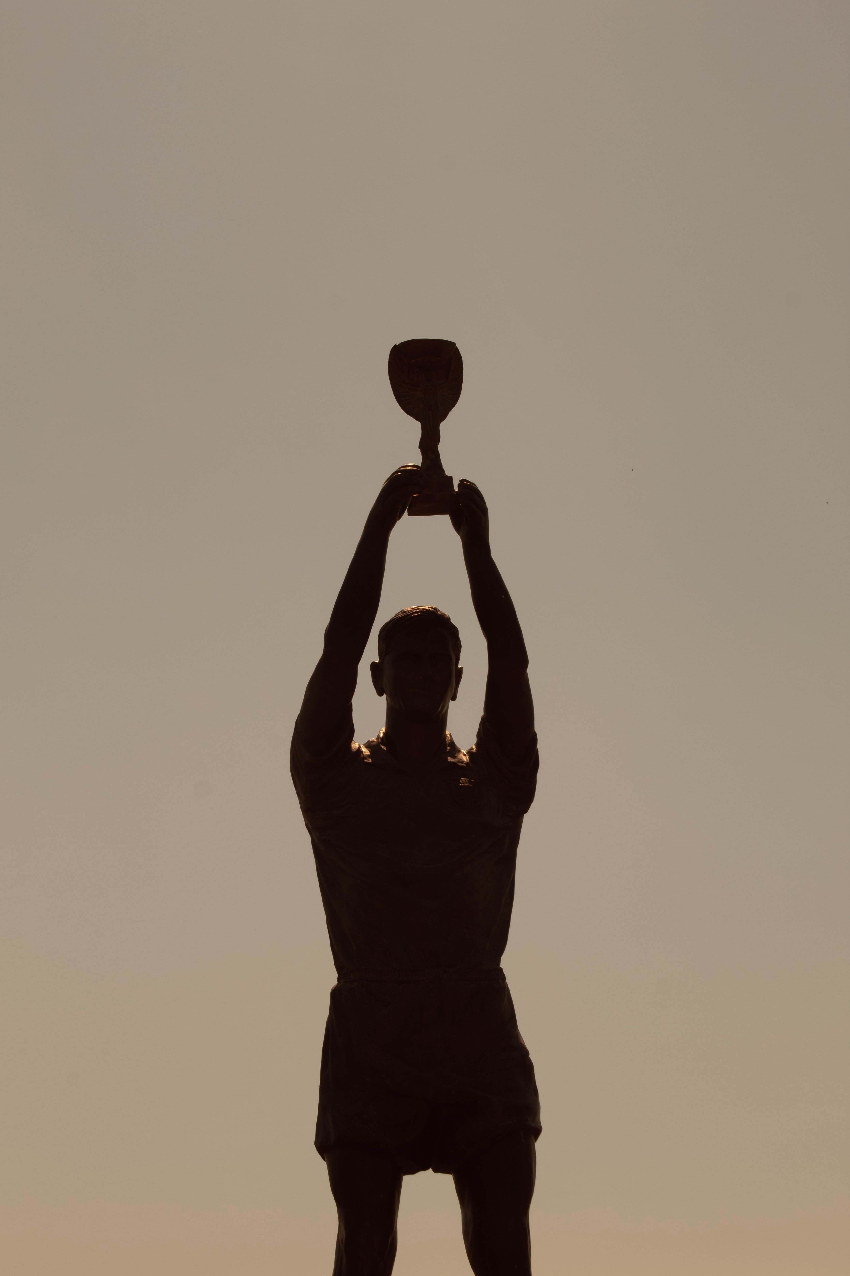 A Silhouetted Statue of a Man Holding a Cup Over Head · Free Stock Photo
