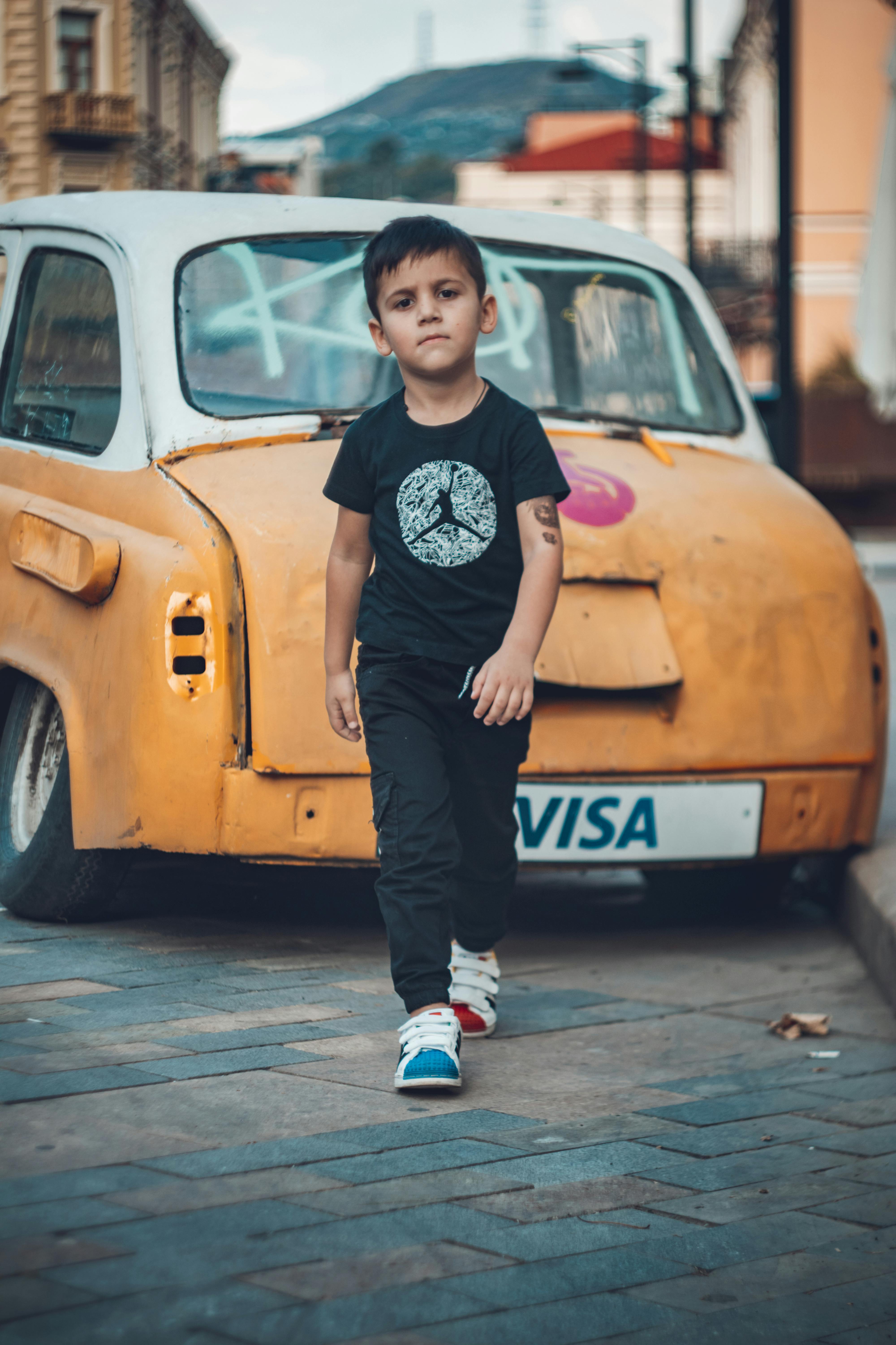 Boy in Black Clothes Walking Beside Yellow Car · Free Stock Photo