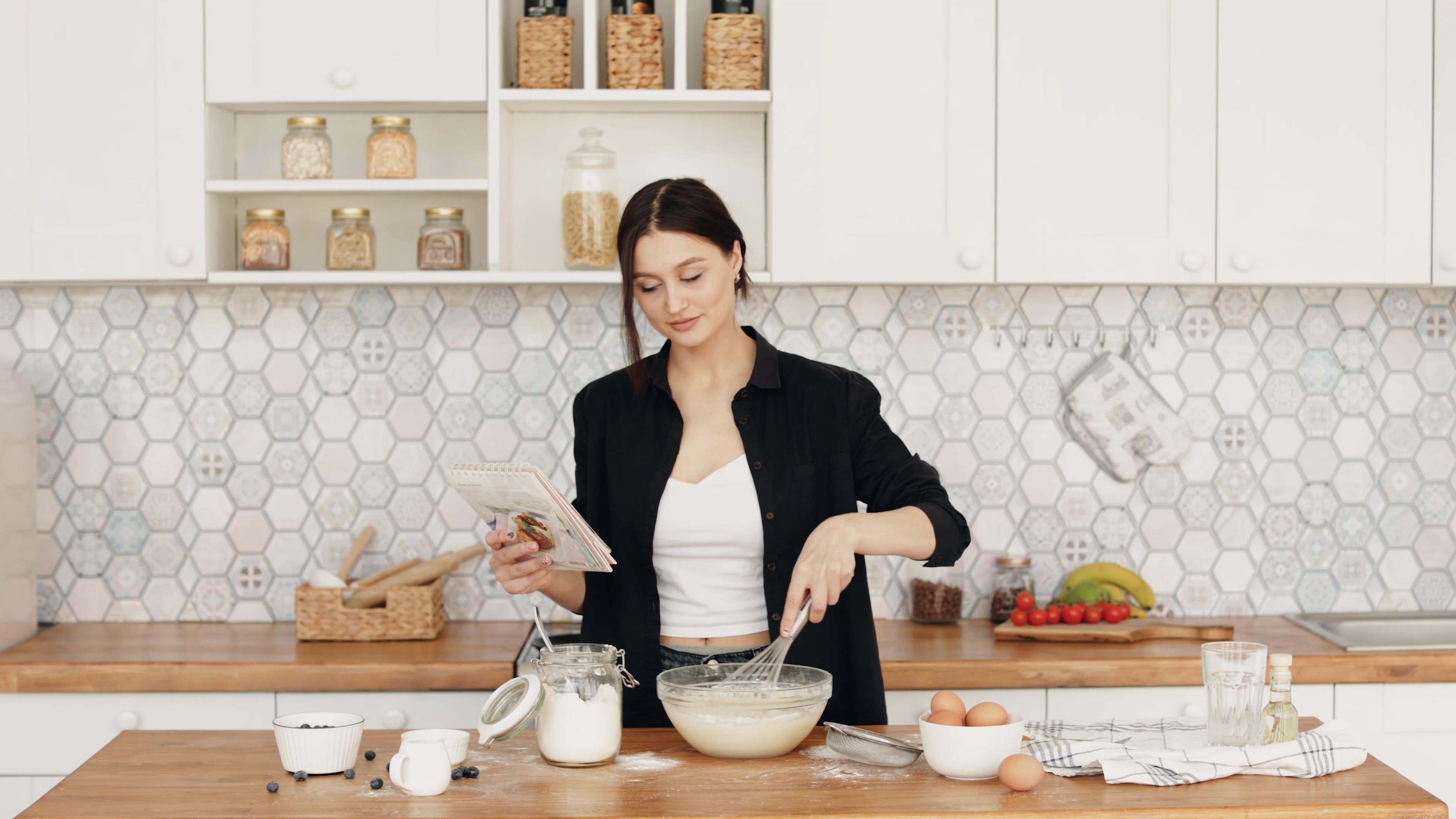 Woman Looking at a Cookbook while Whisking the Flour in a Mixing Bowl ...