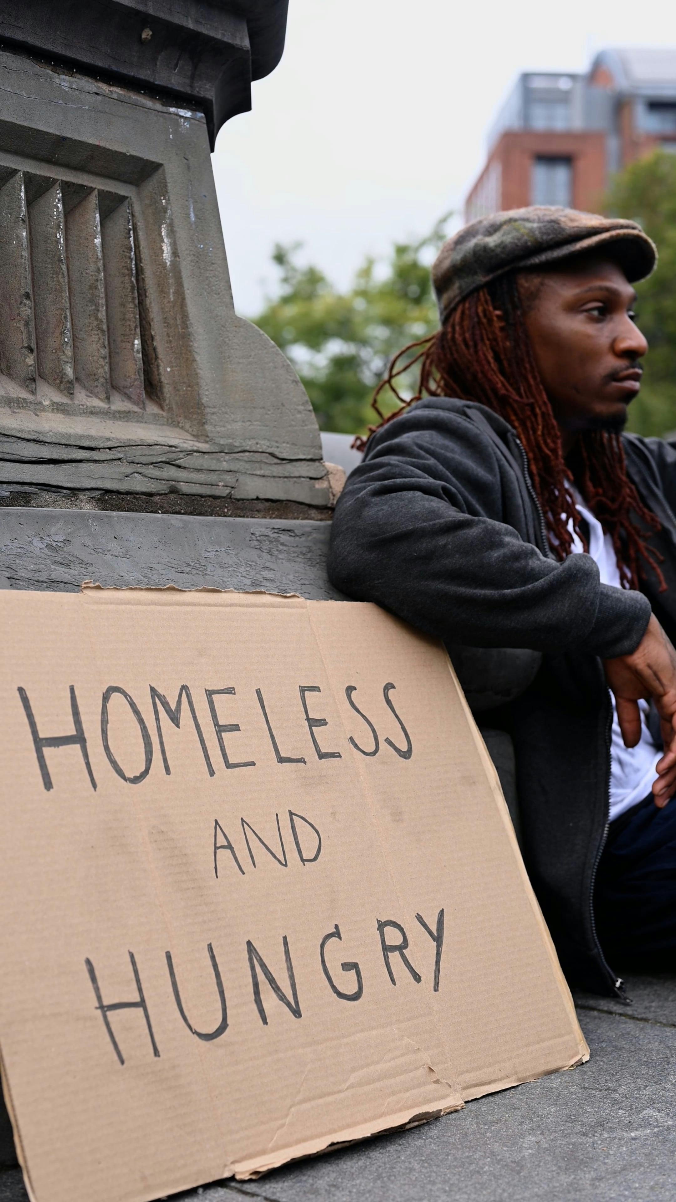 A homeless man sits on a bench with a sign that says homeless and ...